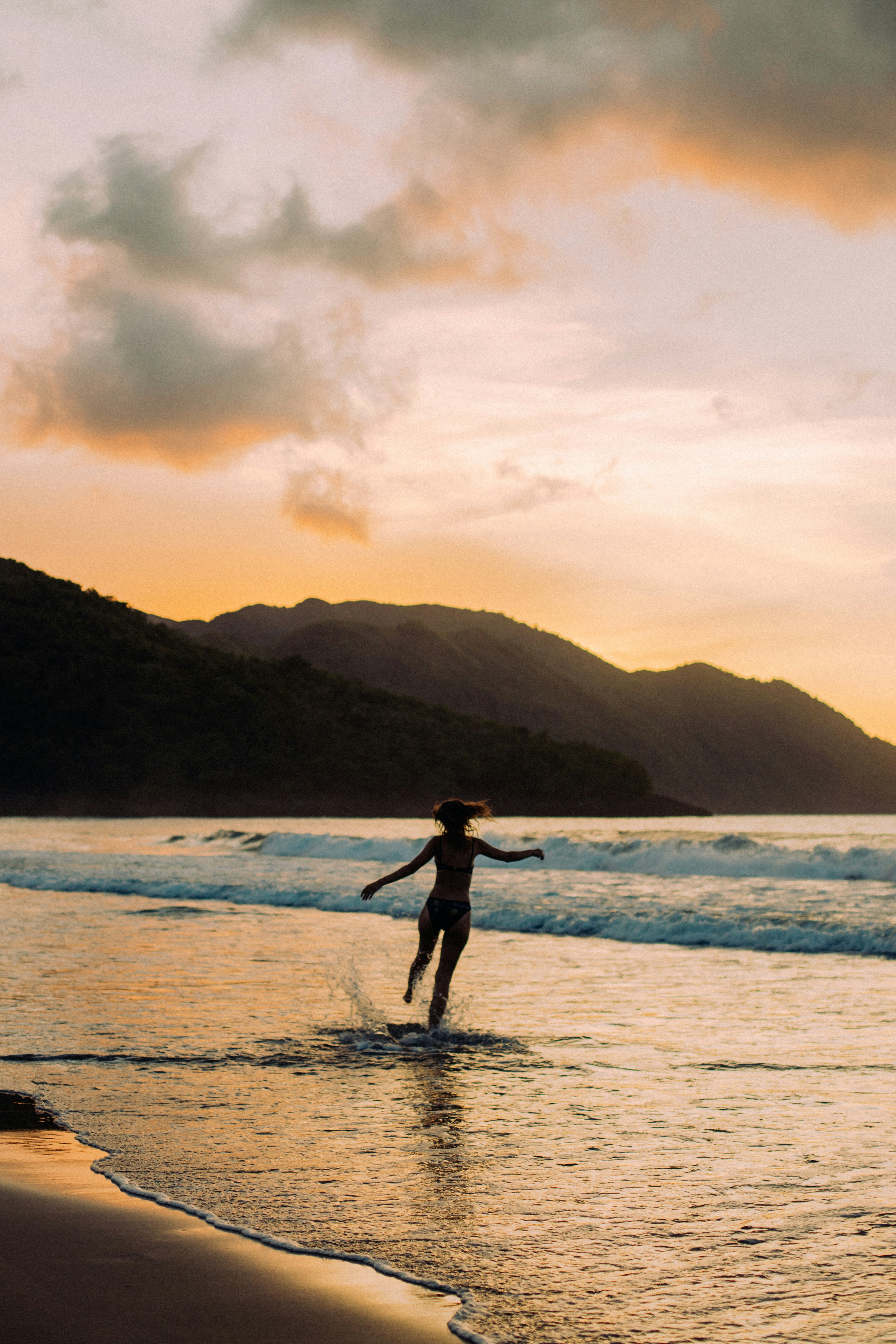 A lone figure skim-surfing at sunset along a tranquil shoreline, silhouetted against the glowing waters and distant mountains—capturing a serene beach moment. The scene appears as a photography print or still from a travel logo shot.