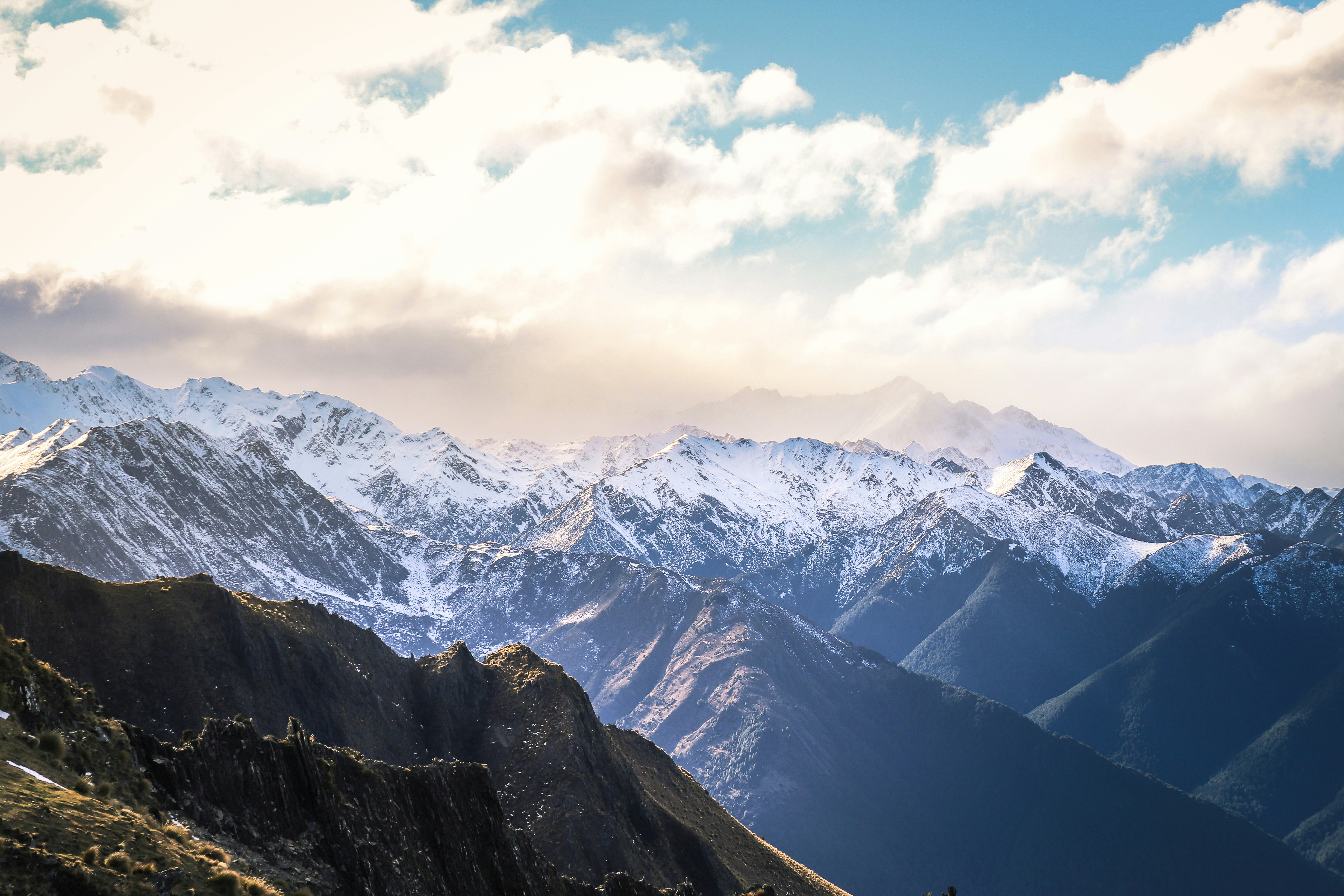 Snow-dusted mountain ridges sweep across the frame under a blue, cloud-swept sky. A deep valley and shadowed slopes provide dramatic depth and texture.
