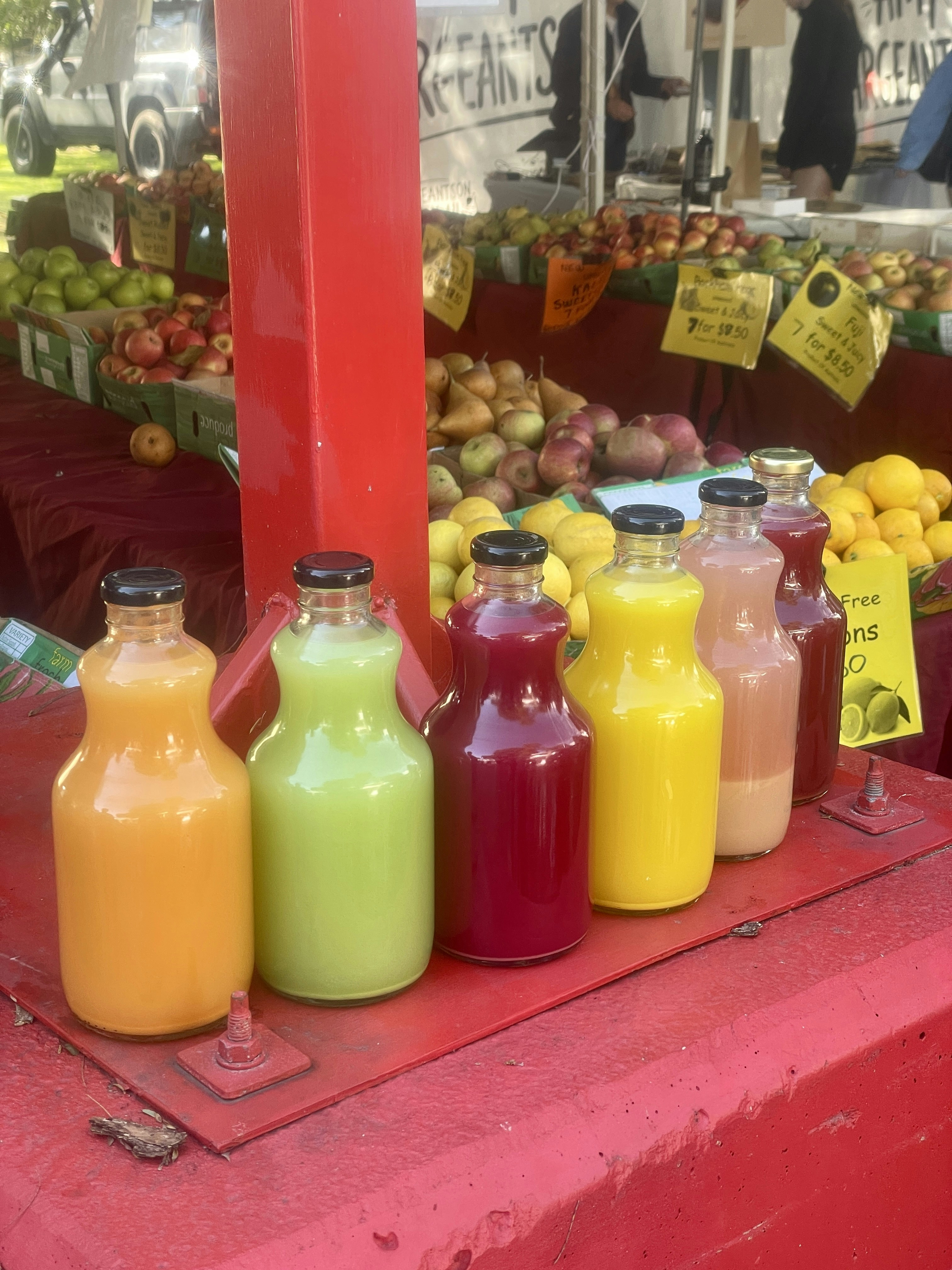 Row of colorful juice bottles on a red market stall with fruit in the background, highlighting a lively produce scene.