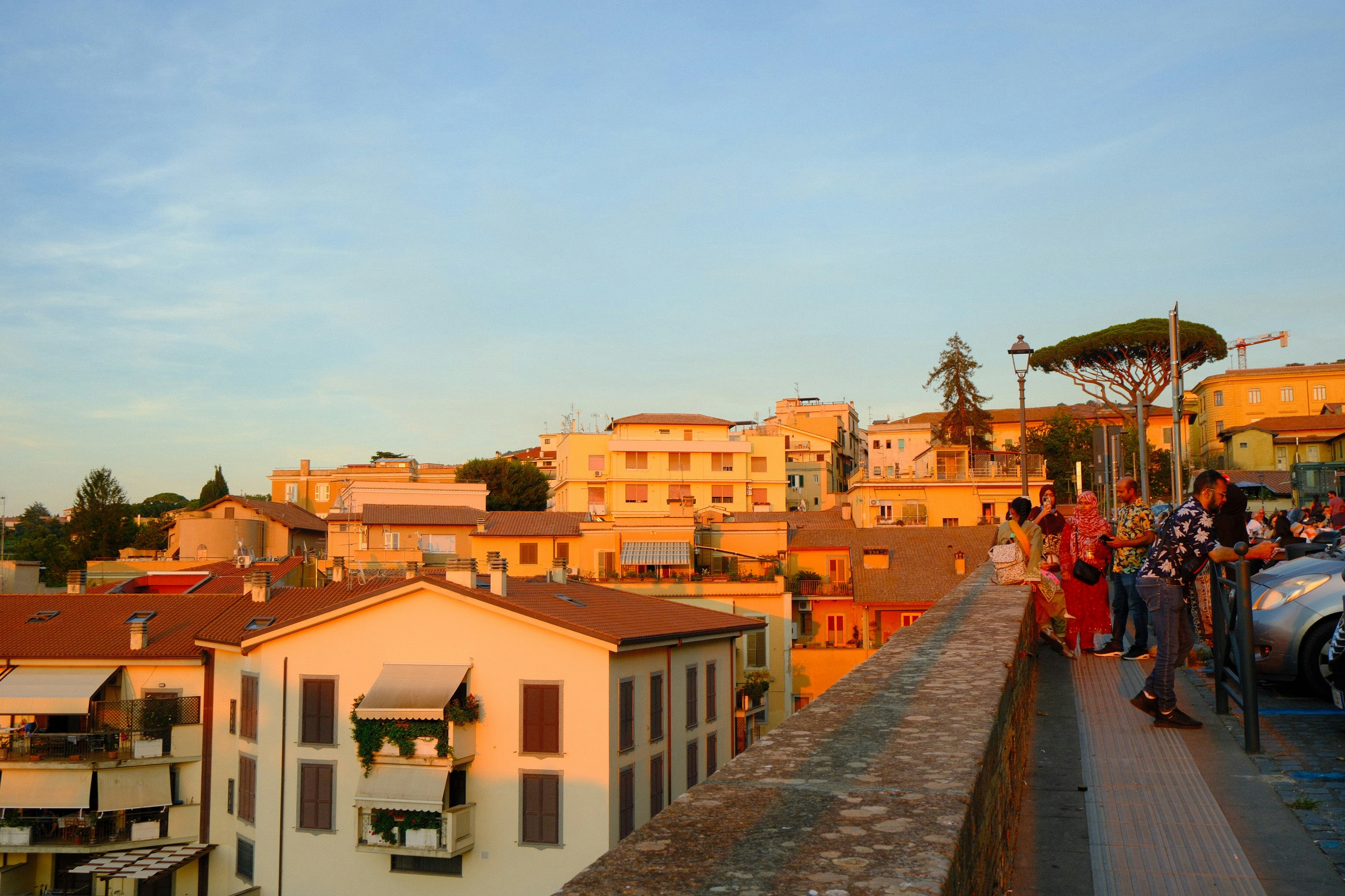Sunlit cityscape with a stone terrace edge and pedestrians along a promenade, bathed in warm evening light; a mix of pastel buildings and parked cars fill the scene.