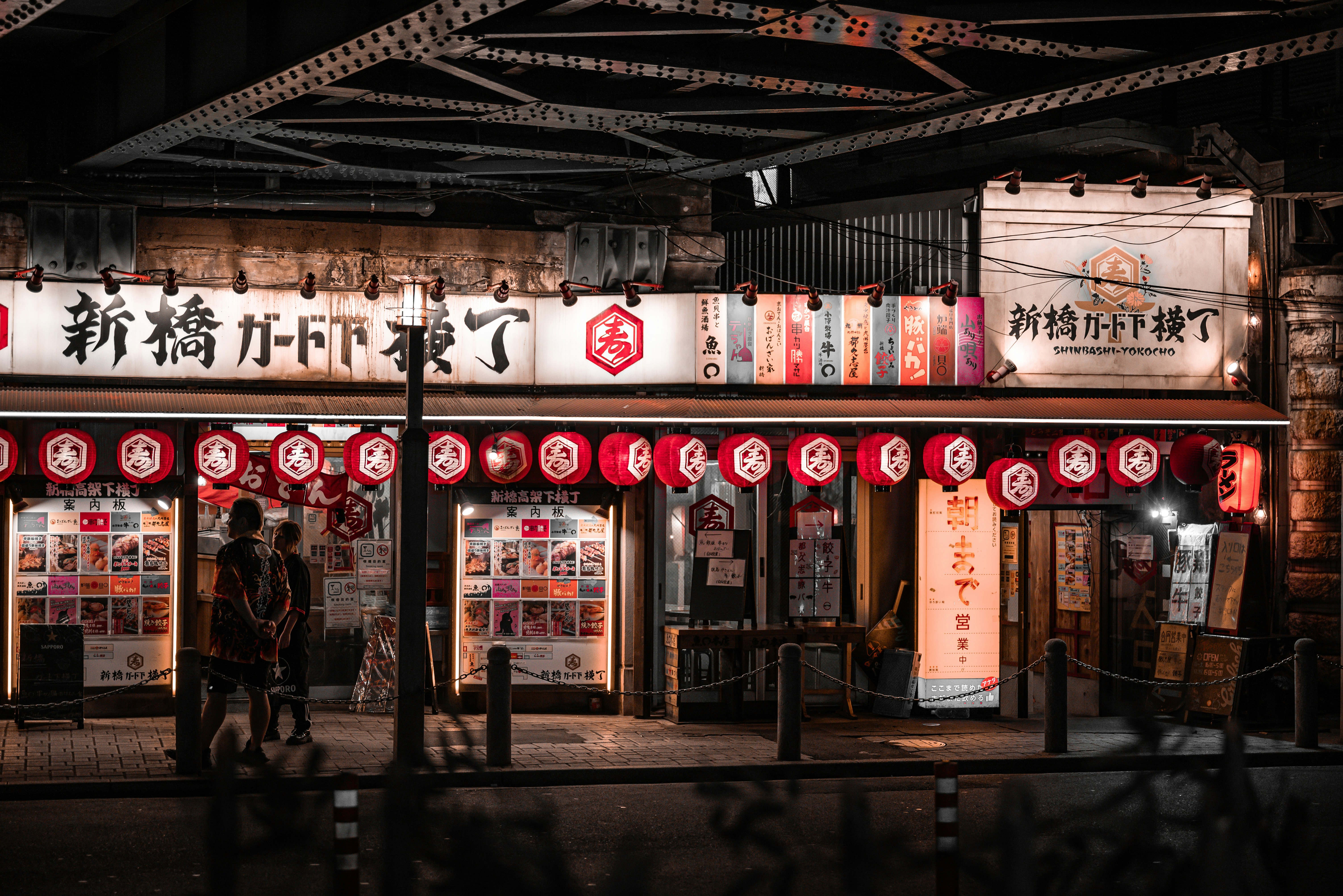 A dimly lit storefront row with red lanterns and signage glows along a quiet street; silhouettes of two pedestrians pass as warm light spills onto the pavement. The scene captures a nocturnal urban vibe with a traditional aesthetic.