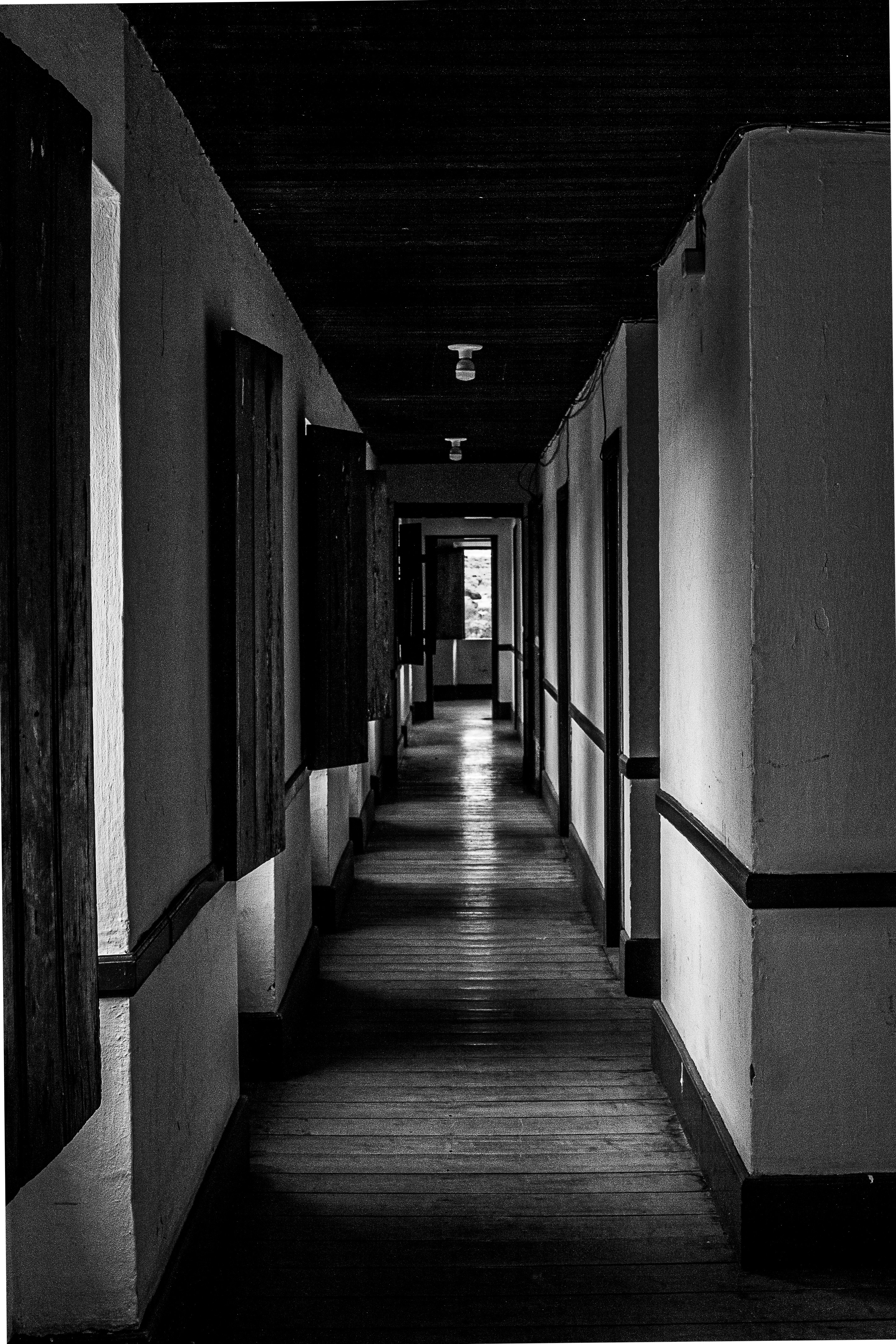 Dark, monochrome hallway with a bright doorway at the far end; the composition emphasizes perspective and leading lines through the centered corridor.
