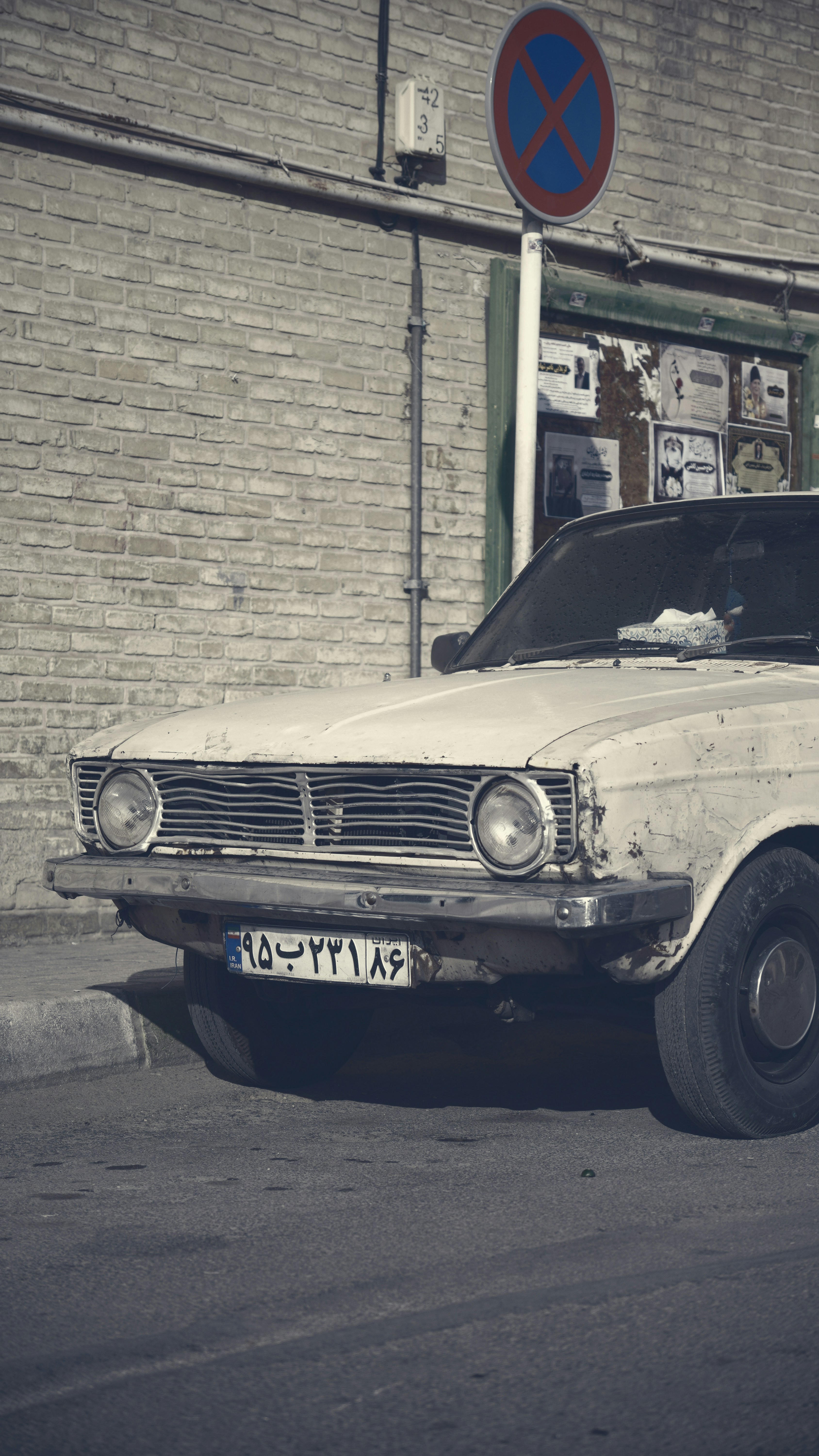 A weathered vintage car sits parked along a brick wall beneath a round traffic sign, with posters on a bulletin board in the background.