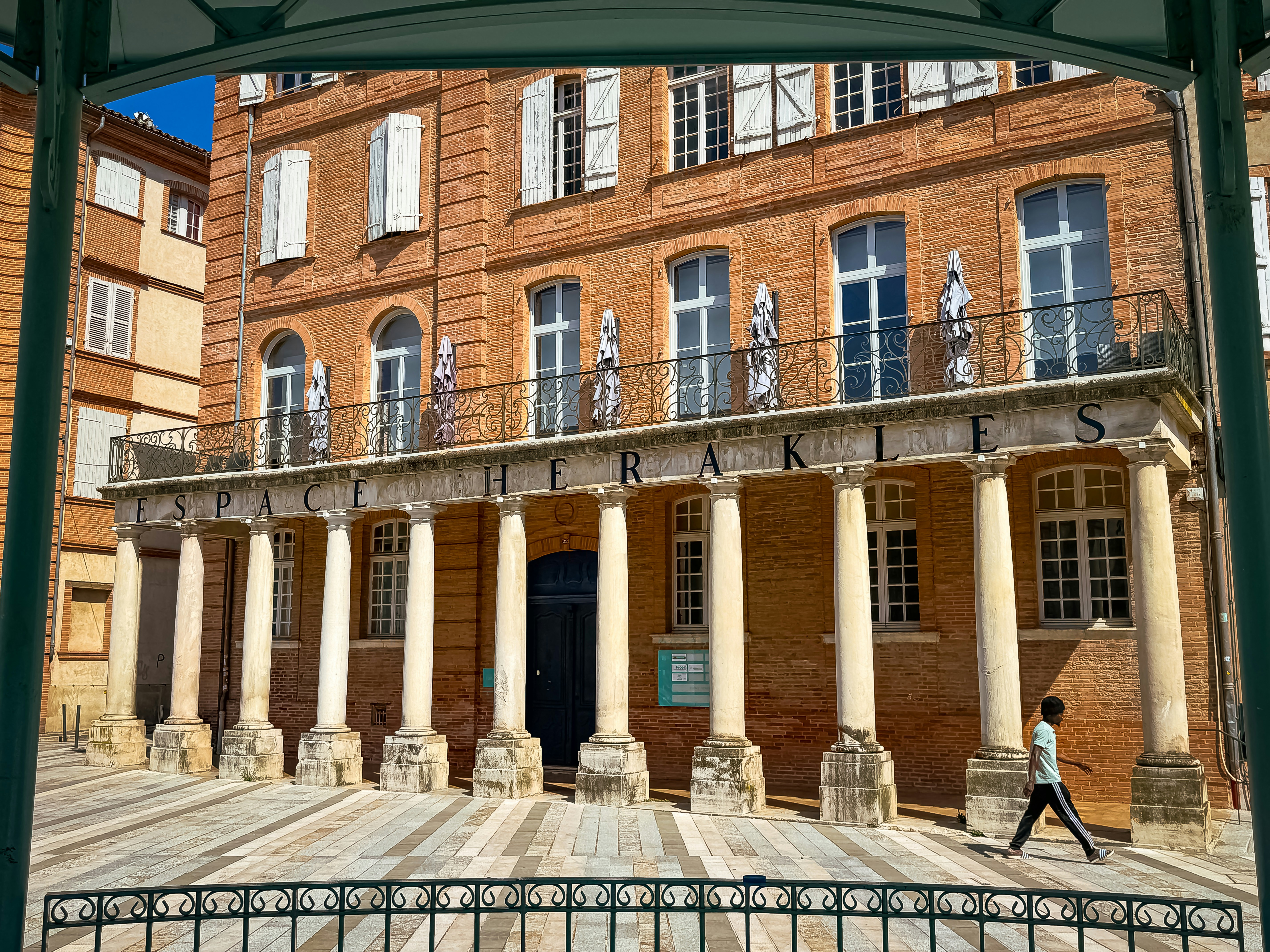 Sunlit brick building with an arched colonnade and stone columns dominates the scene; a lone passerby crosses a tiled courtyard beneath a shaded canopy.