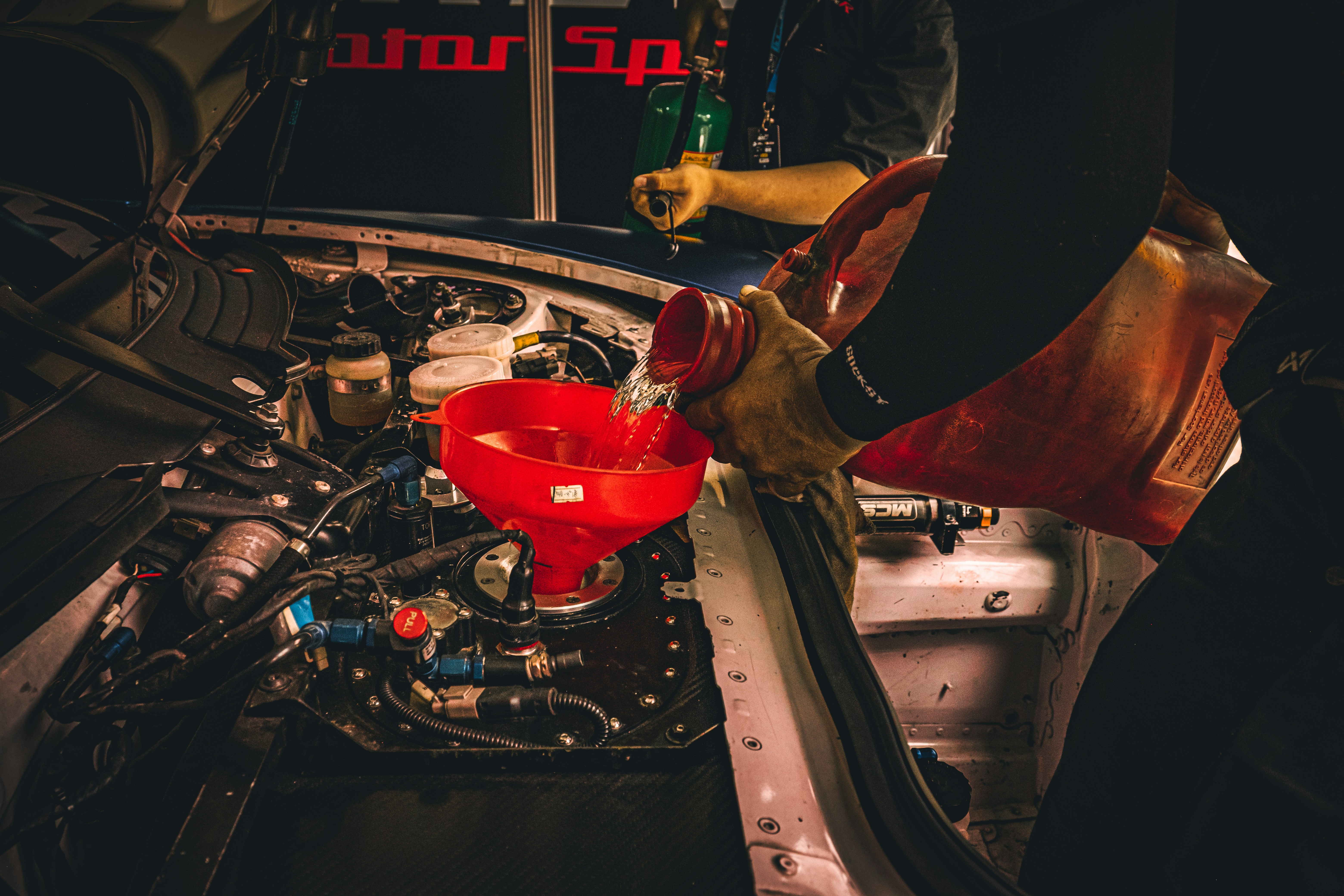 Engine Bay Ritual: Pouring Emission into a Red Funnel in a Race Car Garage