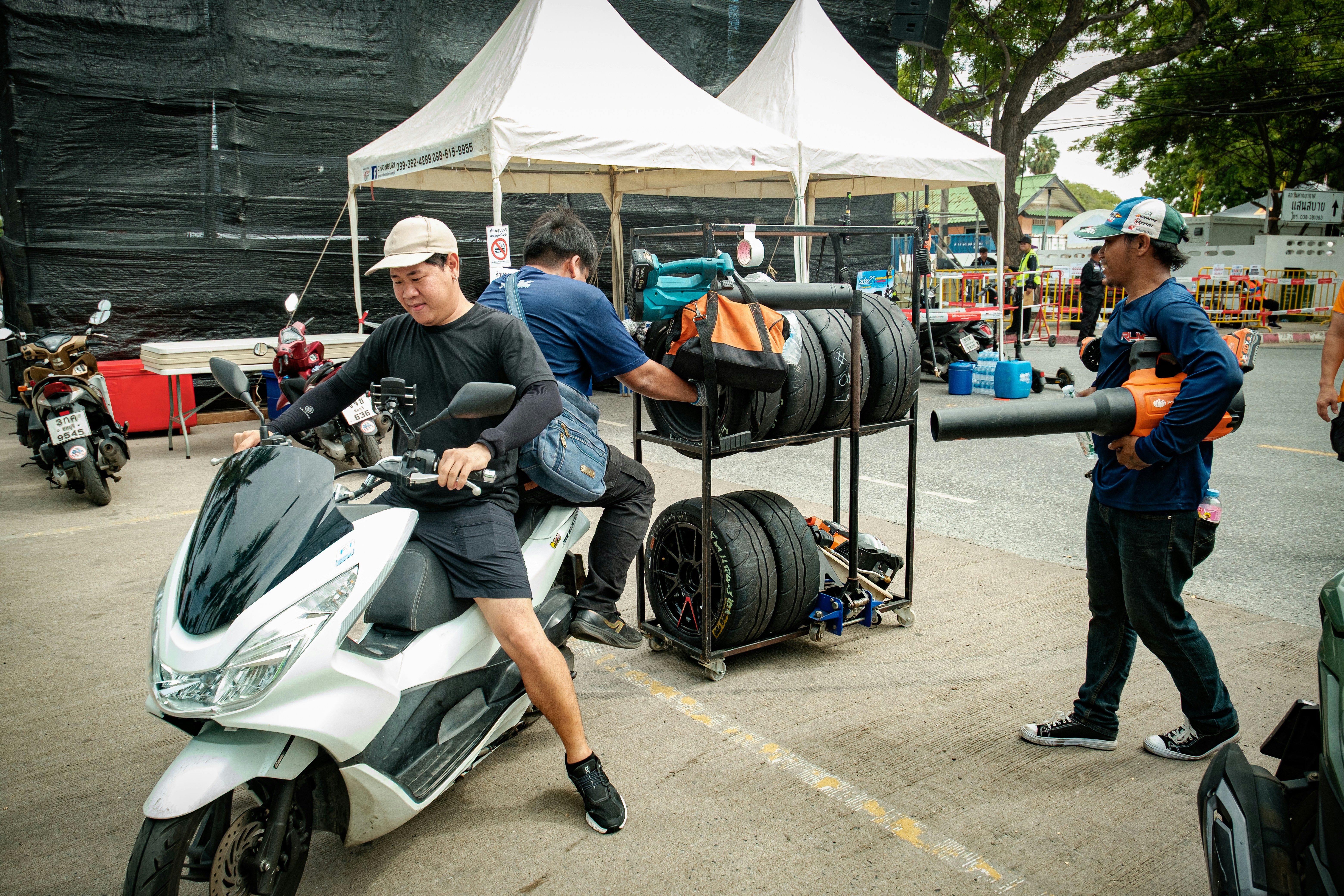 A candid, resourceful behind-the-scenes shot of race team crew members utilizing a white scooter to tow a rolling rack loaded with race tires and power tools, while another mechanic walks alongside carrying a leaf blower for track cleaning. Captured in the pit paddock area during the 2026 Bangsaen Grand Prix / Thailand Super Series at the Bangsaen Street Circuit in Chonburi, Thailand. This image highlights the creative problem-solving, busy logistics, trackside preparation, and authentic local flavor of Asian motorsport events.