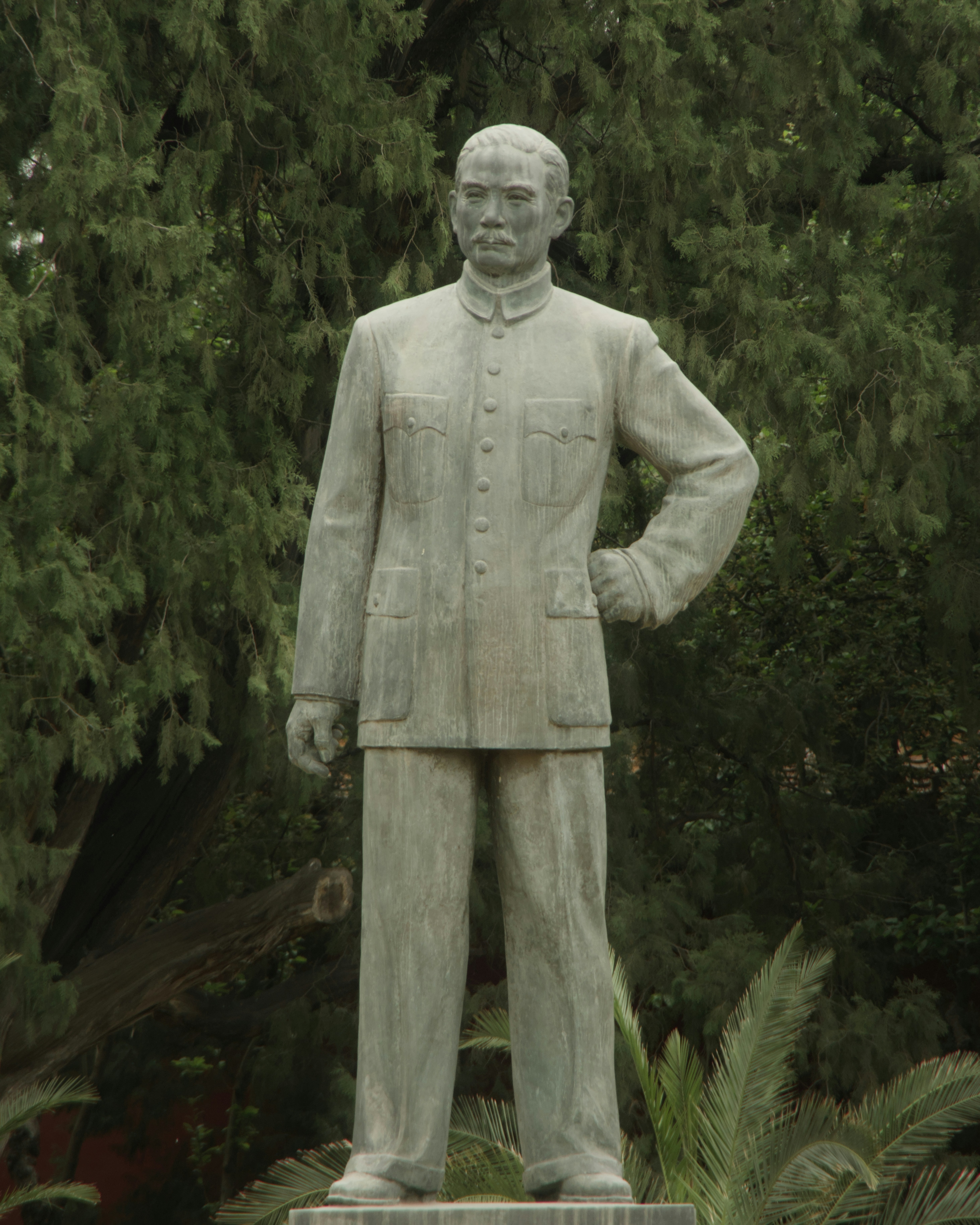 Stone statue of a stern, uniformed man stands with one hand on hip among greenery; a solemn monument captured in soft daylight. This is a sculpture, not a living subject.