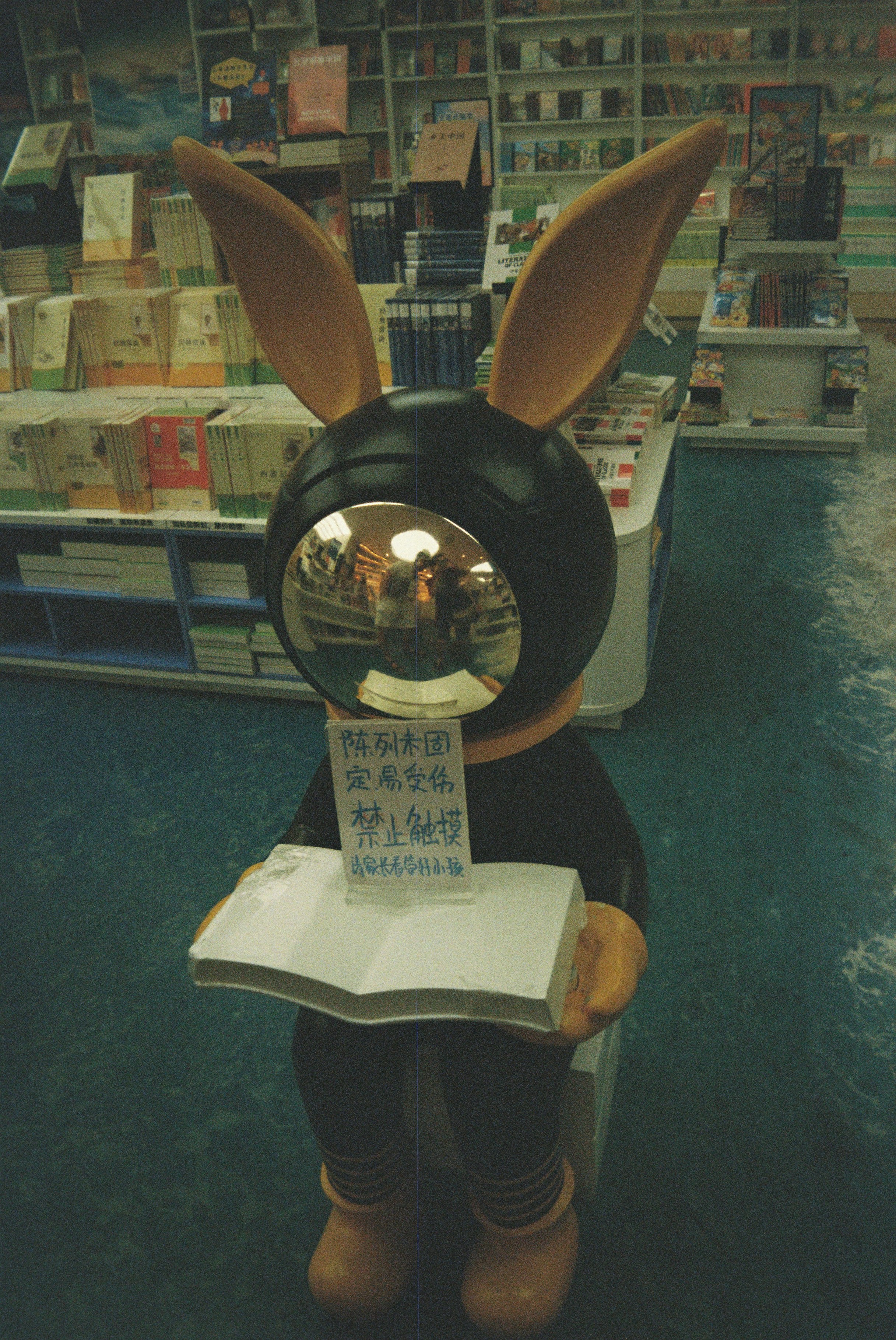 A life-sized rabbit-like mascot sits holding a book inside a bookstore in a shopping mall in Sanya, Hainan Island, China. Instead of a face, the mascot features a reflective mirror surface that captures the photographer and their friend standing in front of it. The dark, slightly gloomy lighting creates a mysterious and surreal atmosphere, making this an introspective and dreamlike portrait.