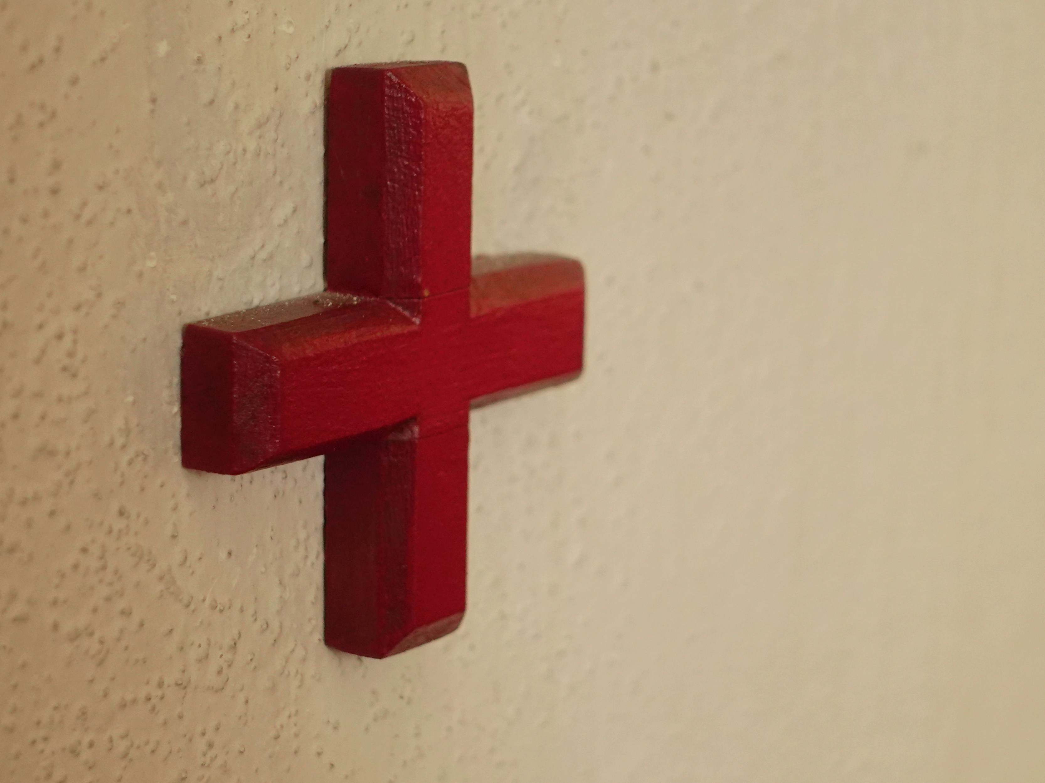 A small red cross protrudes from a light-textured wall, casting subtle shadows and providing a tactile, geometric focal point.