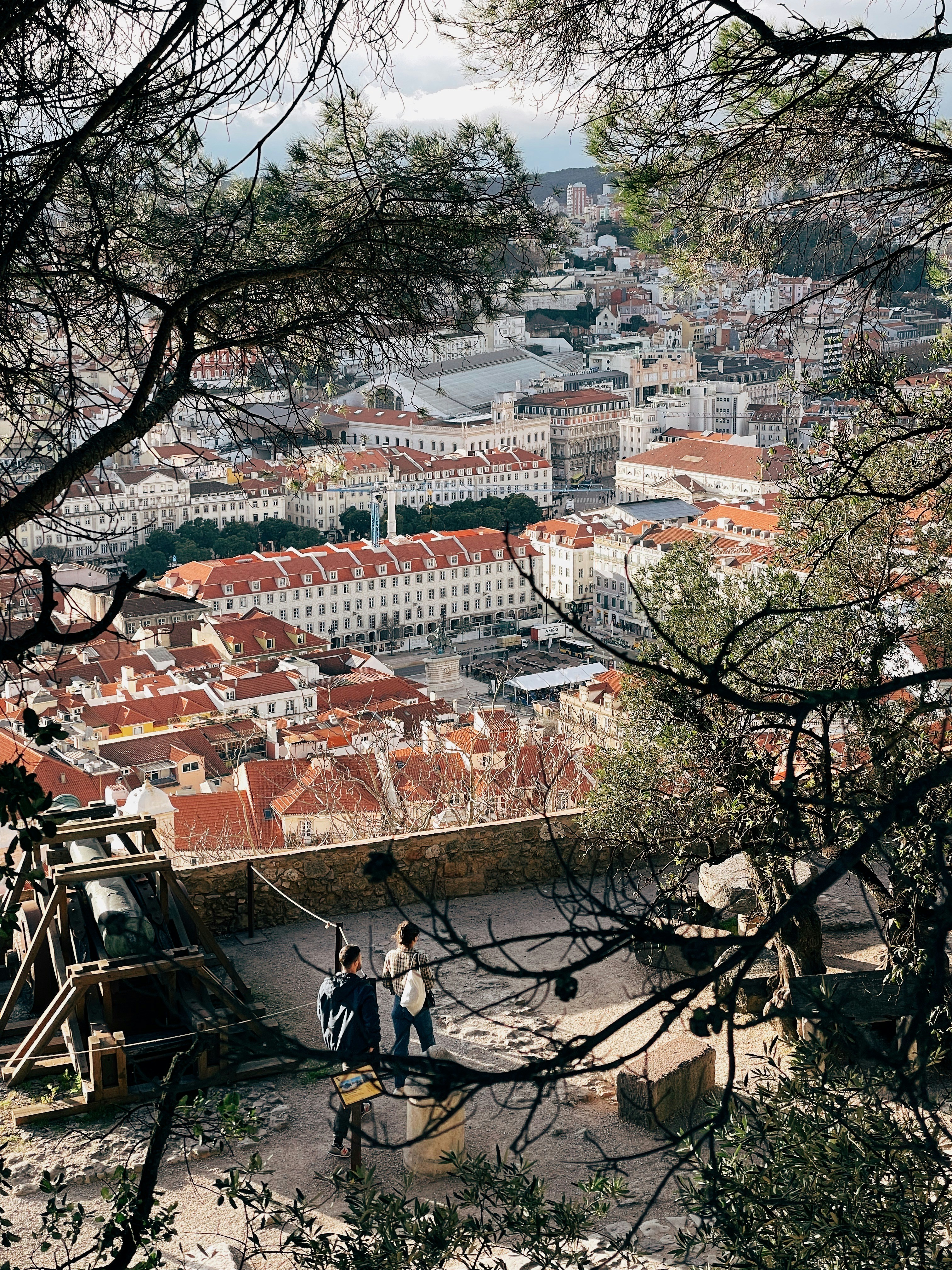 Elevated view of a European town with terracotta roofs framed by tree branches; a small viewing area and two people foreground the skyline.