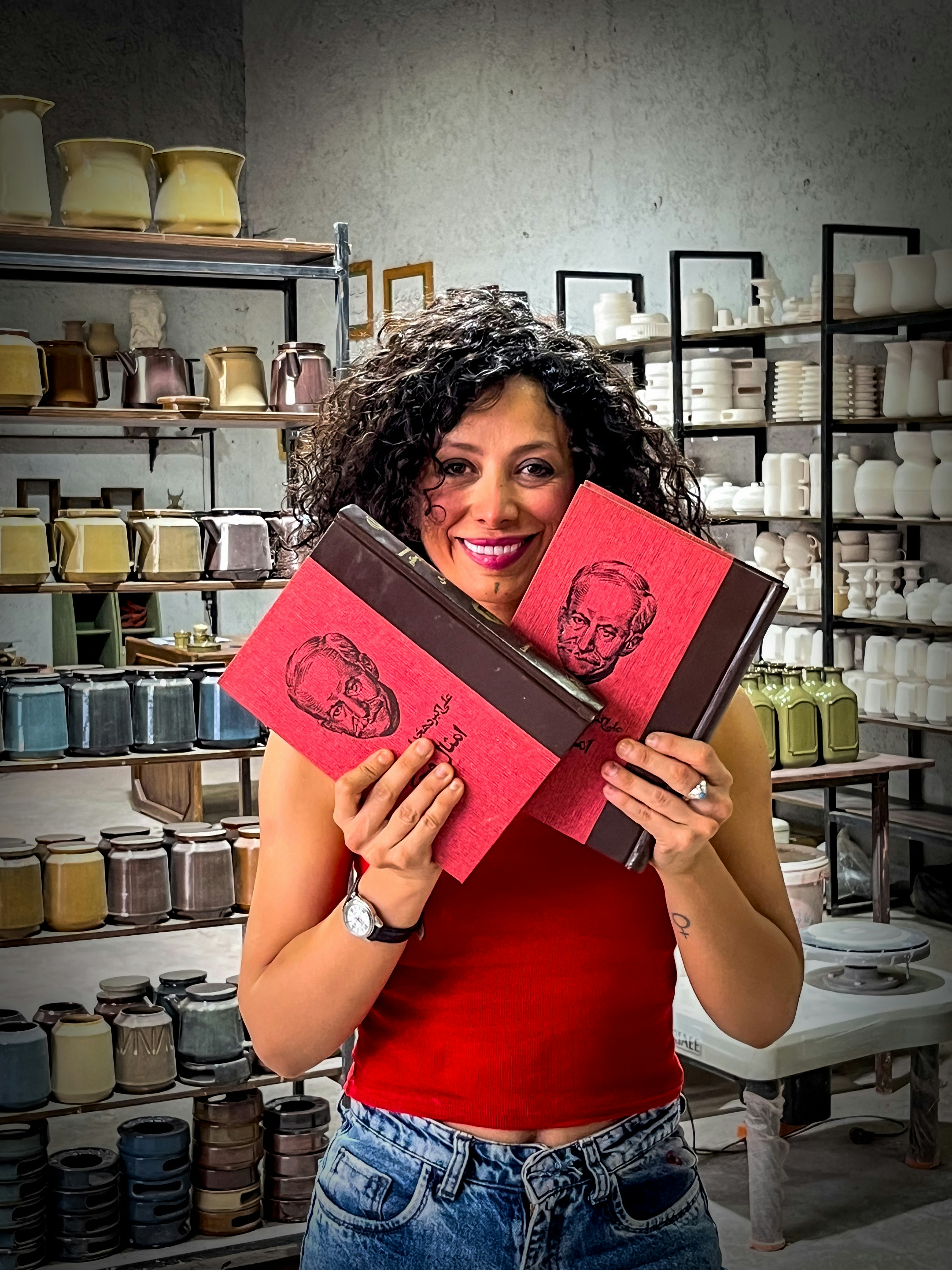 Vibrant portrait of a woman in a red top holding two red sketchbooks in a ceramic studio; colorful shelves of pottery surround her in the background.