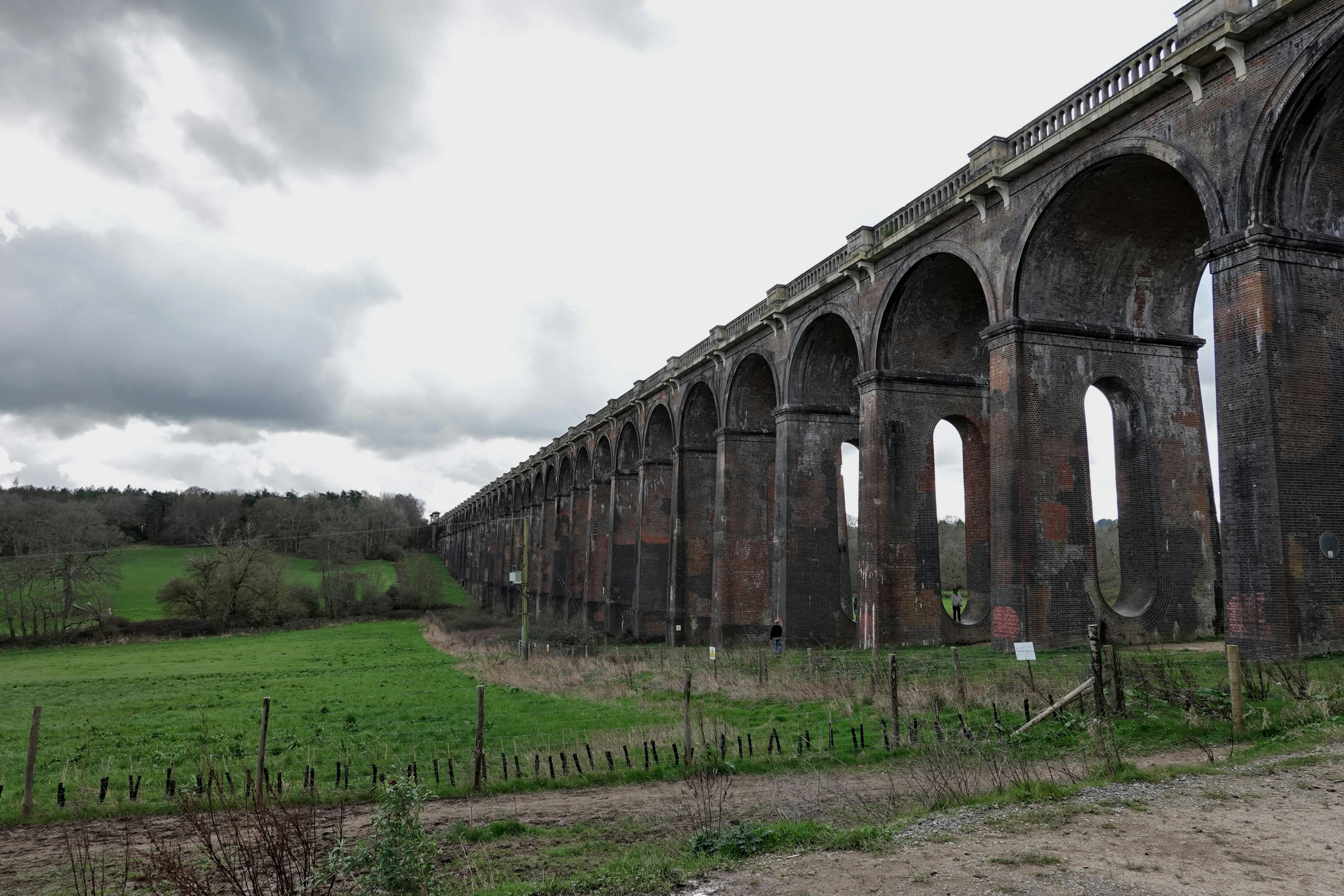 Brick arches of a long viaduct rise above a grassy landscape with a dirt path in the foreground.