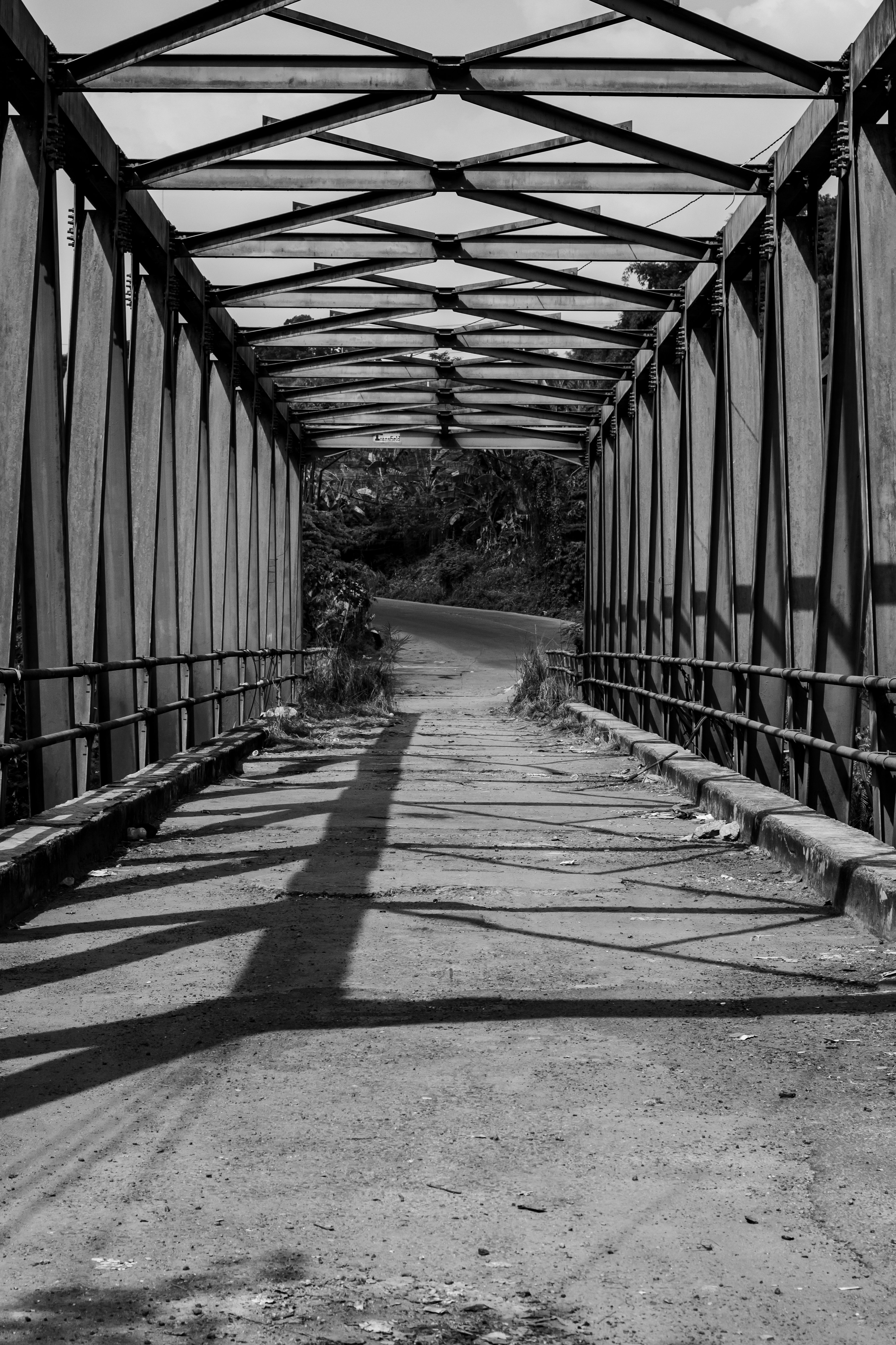 Monochrome image of an empty steel truss bridge with strong geometric shadows along a straight, narrow roadway leading toward a distant bend.