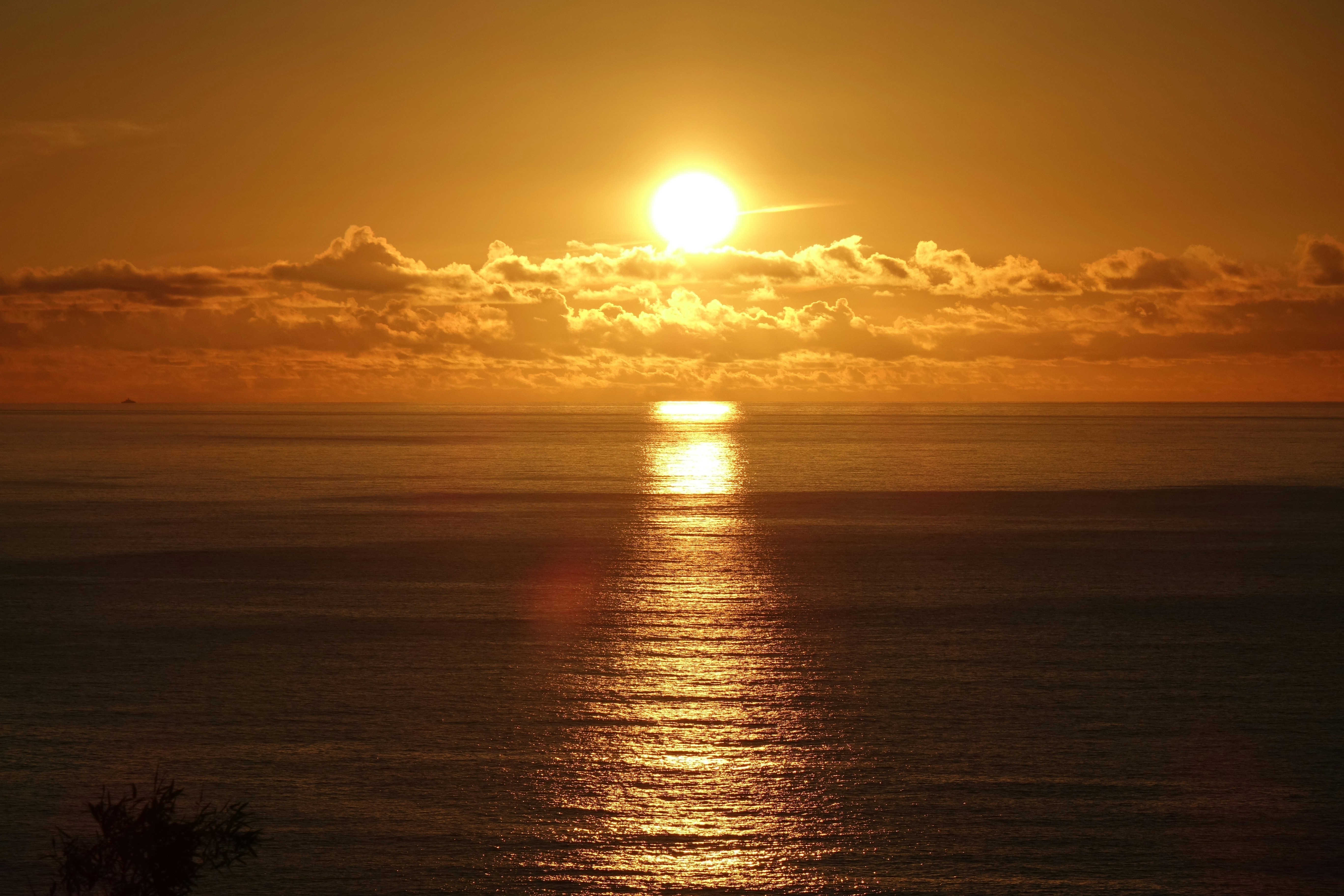 Soft, warm sunset over calm seas with the sun low on the horizon and its reflection rippling toward the shore. The image emphasizes tranquil coastline and atmospheric colors without any human subjects.