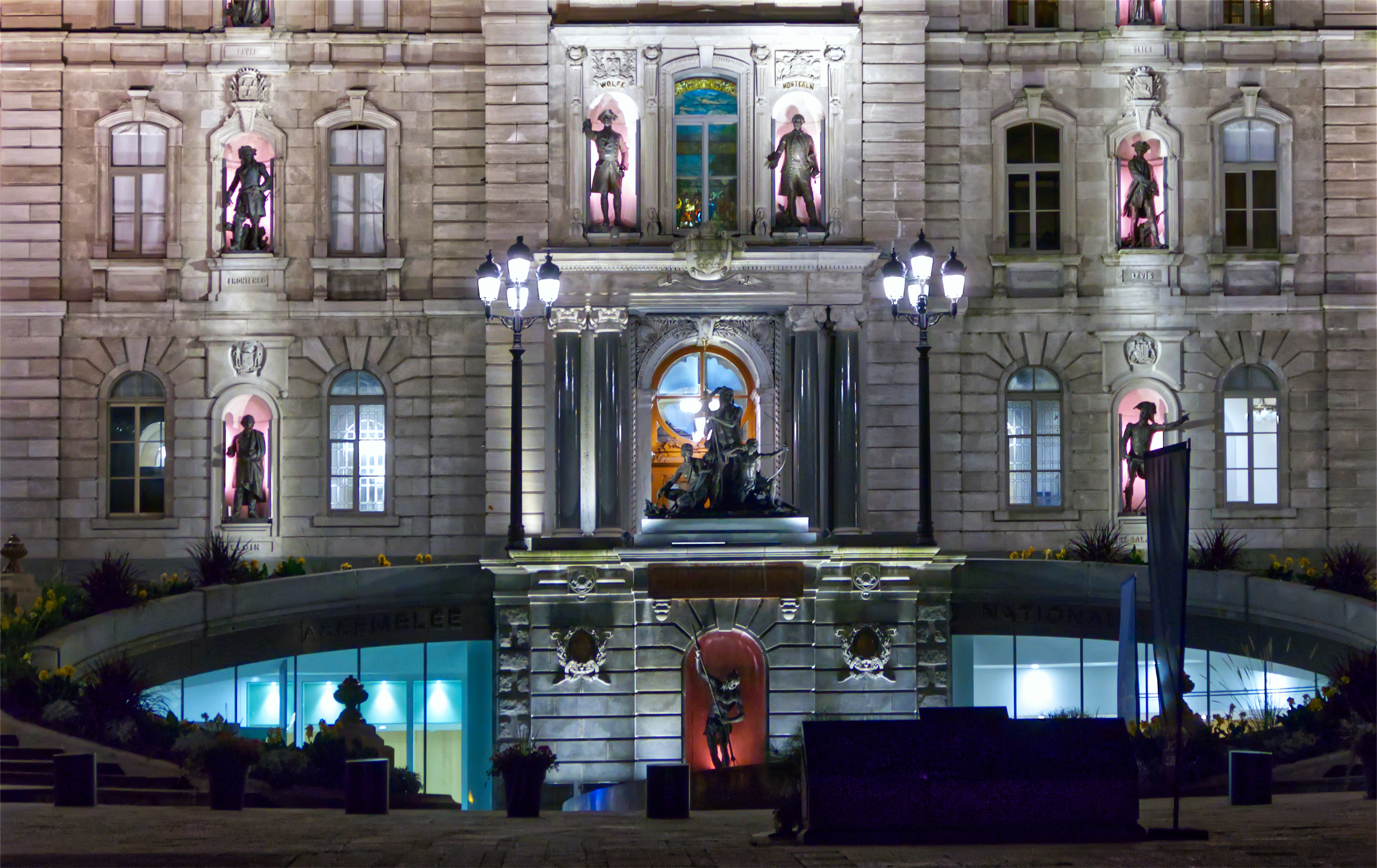 Vue nocturne sur une partie de la façade de l’hôtel du Parlement du Québec. Au Centre, nous apercevons les sculptures allégoriques du « pêcheur à la nigogue » et de la « Halte dans la forêt ». De chaque côté de cette partie centrale, des statues de bronze représentant des personnages marquants de l’histoire du Québec et de l’Amérique française.