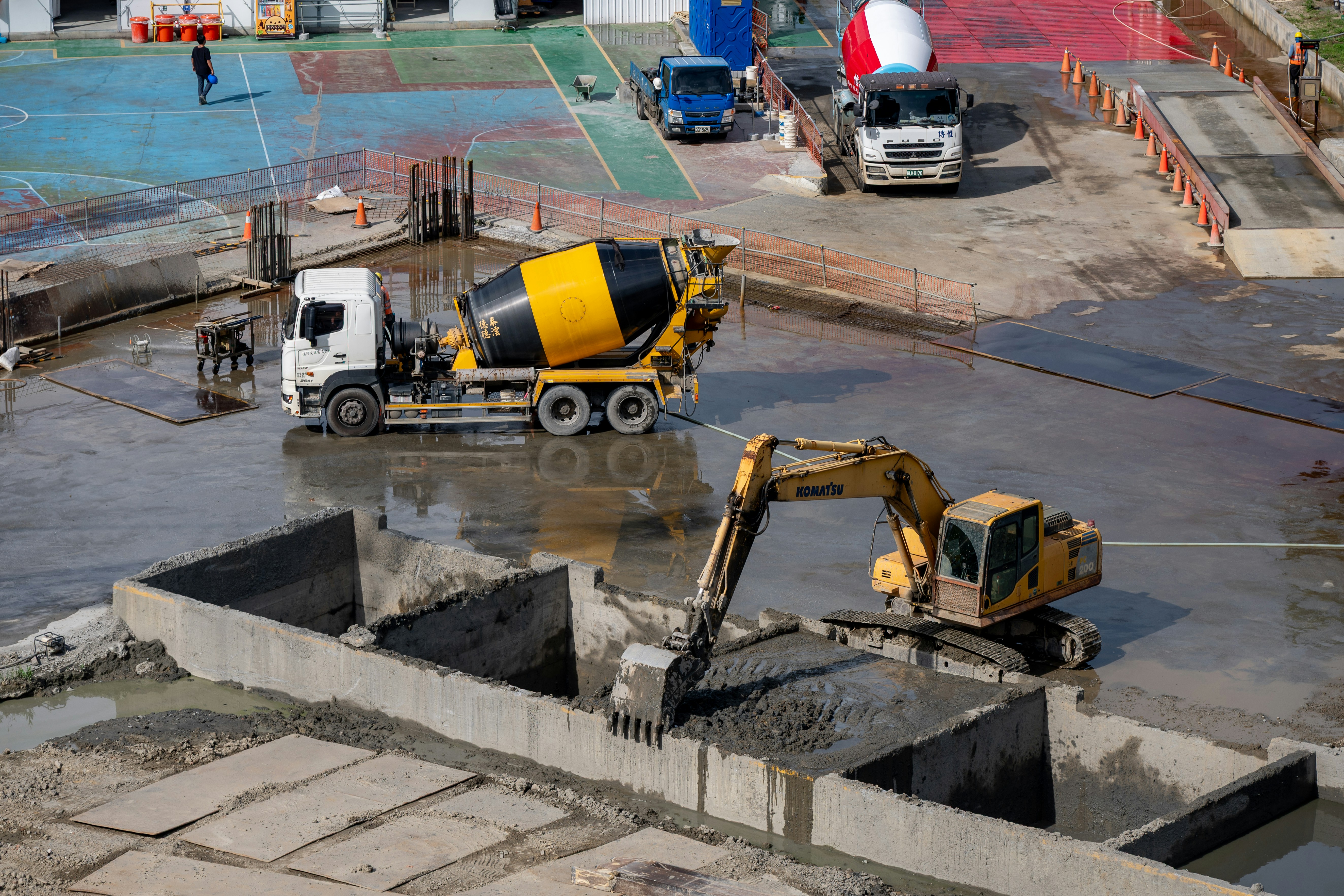 Construction site with a cement mixer and an excavator actively shaping a concrete foundation in a wet, sunlit yard.