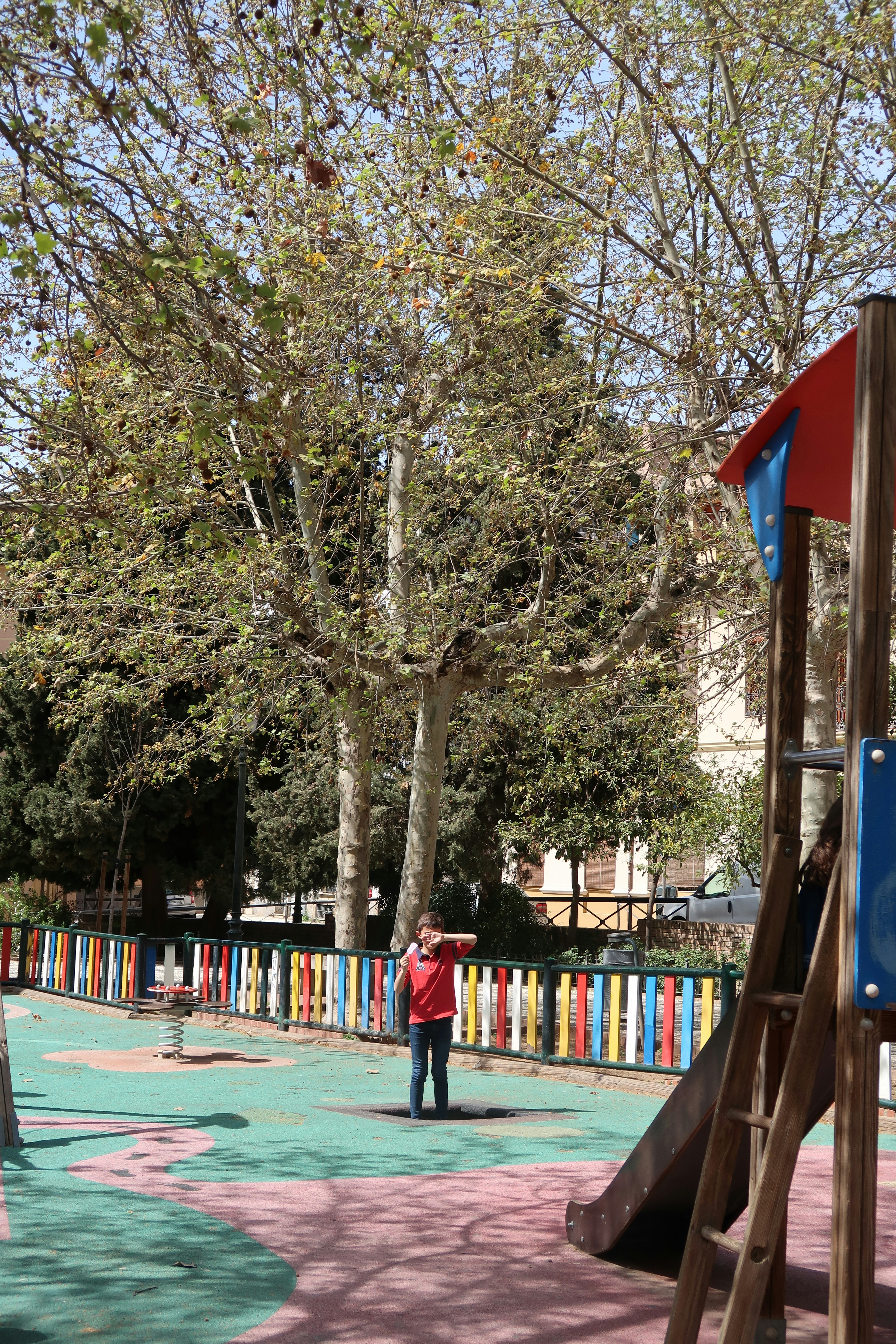 Child in red shirt under a canopy of trees, playground edge in a sunlit park