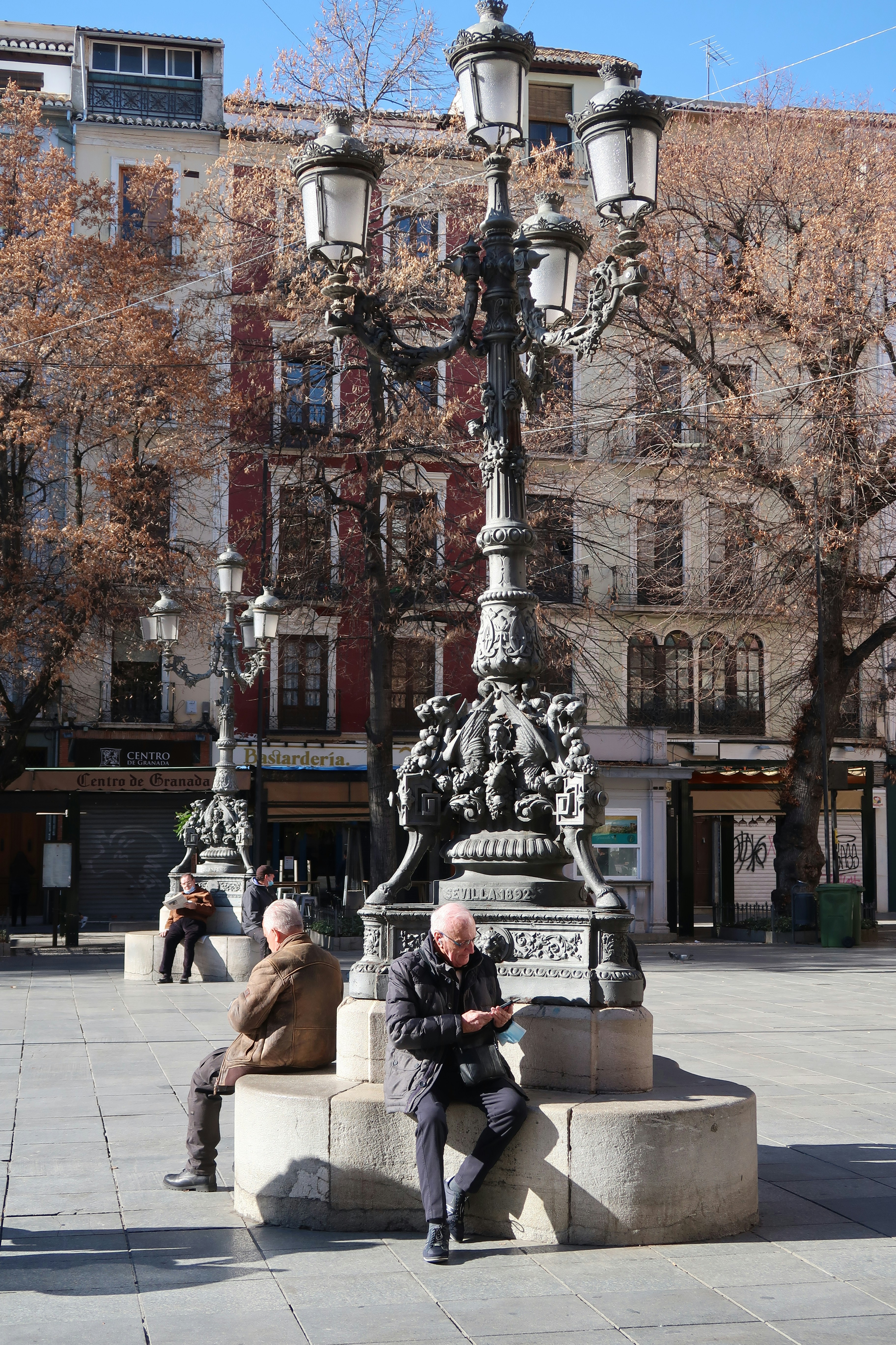 Two seated urban pedestrians at a decorative lamppost in a sunlit plaza