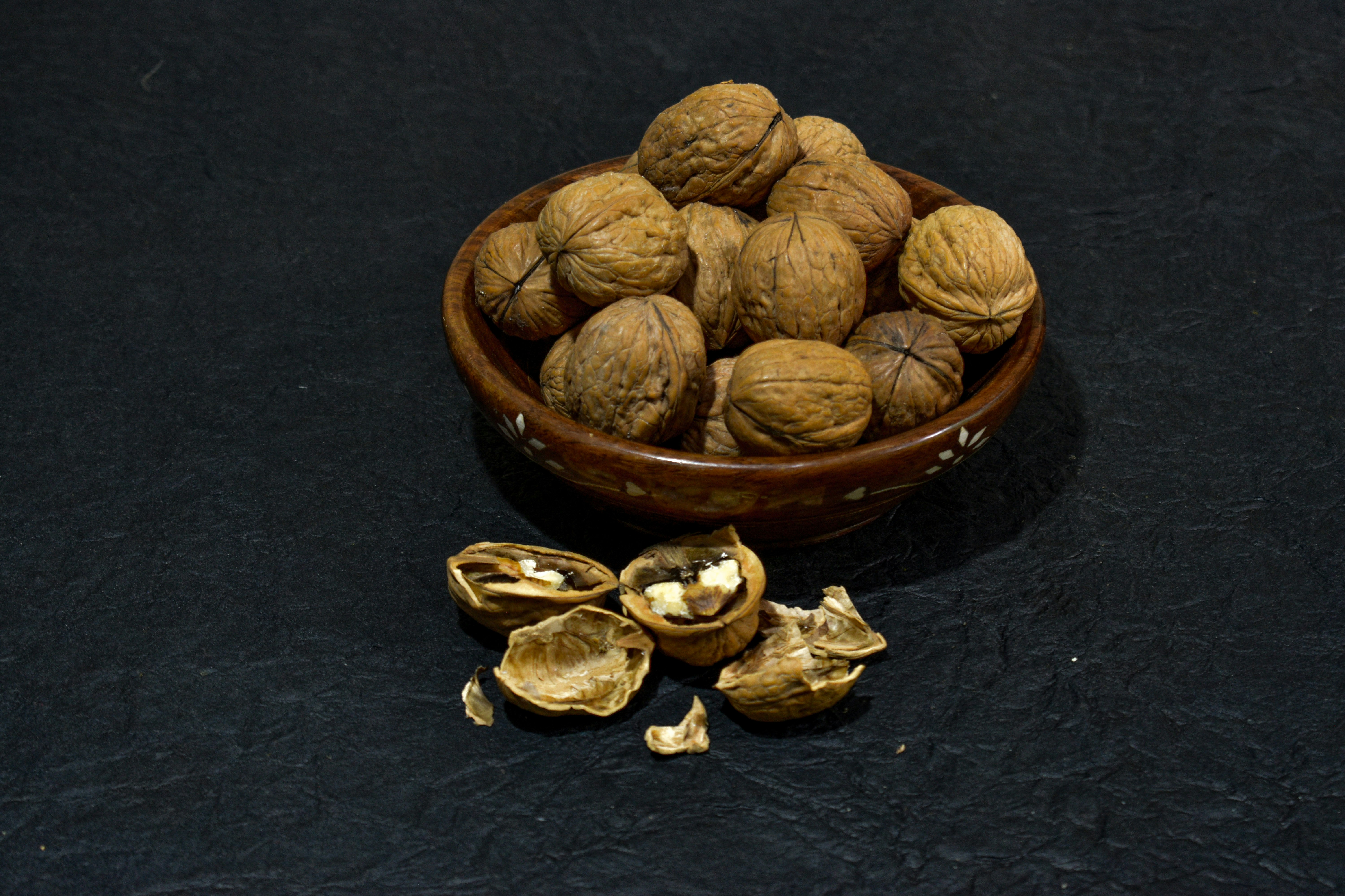 Dark slate backdrop with a wooden bowl of walnuts; some shells cracked open revealing kernels, arranged with a few scattered pieces to the front. A simple still-life emphasizing texture and natural tones.
