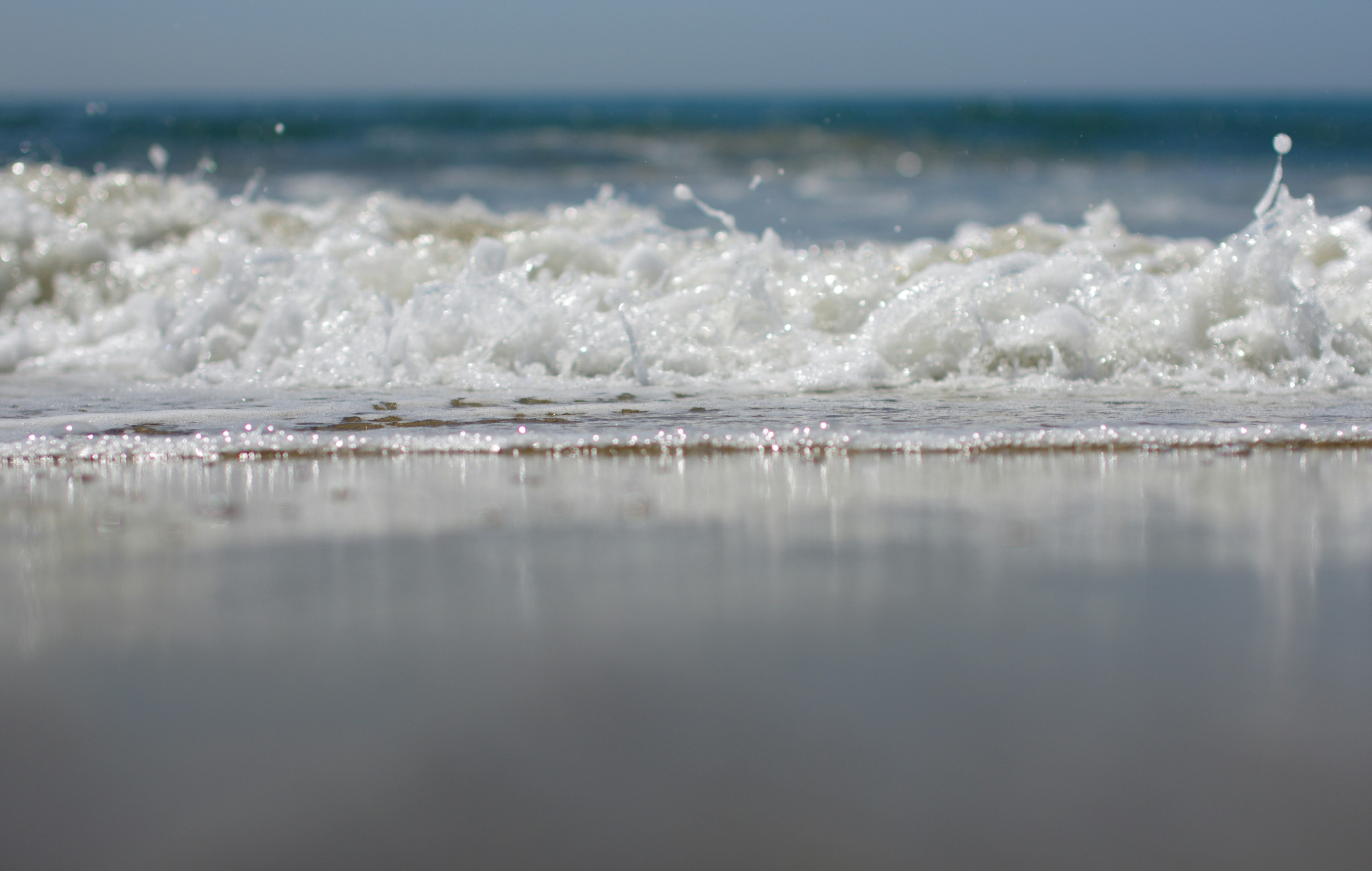 Foamy shoreline with a centered wave crest and distant horizon; minimal foreground with a smooth reflective wet sand, capturing a serene beach moment. (photograph)