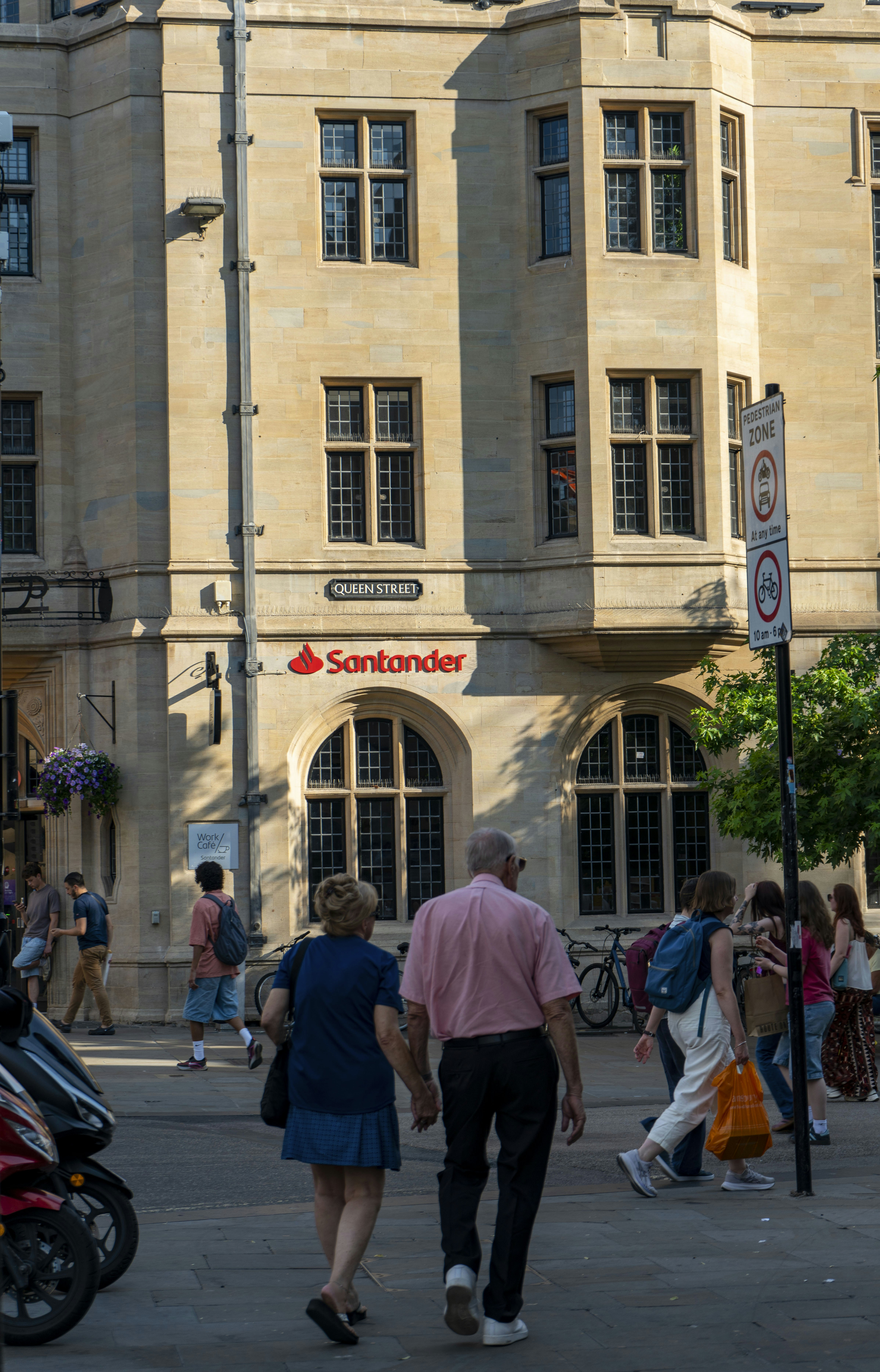 Lively street scene in front of a sandstone building with a Santander sign; pedestrians walk past arched windows and a storefront, captured in daylight.
