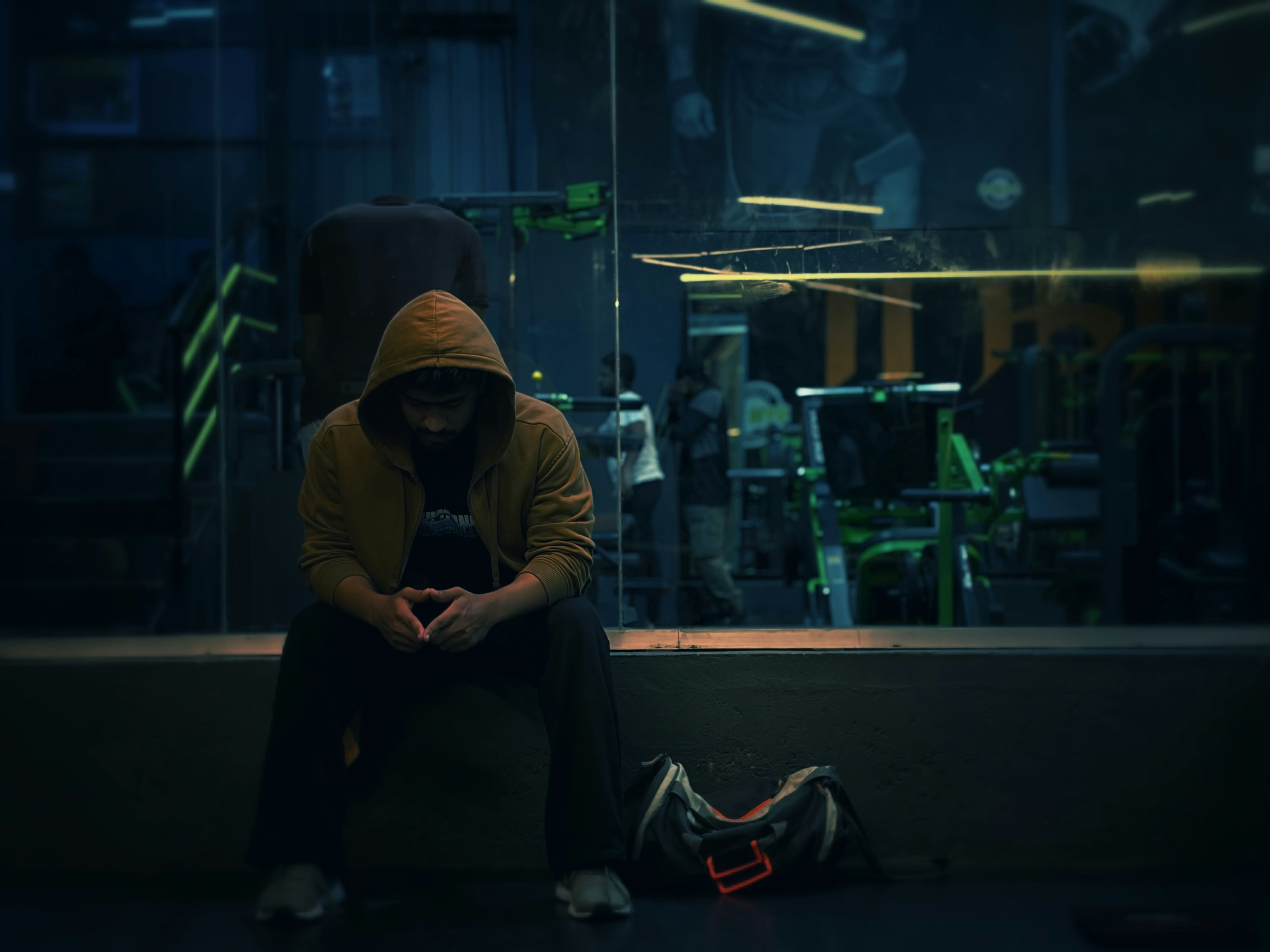 A solitary figure in a yellow hoodie sits on a ledge, head bowed, in a dim workshop seen through a glass wall; a bag rests nearby, and faint neon reflections illuminate the space.