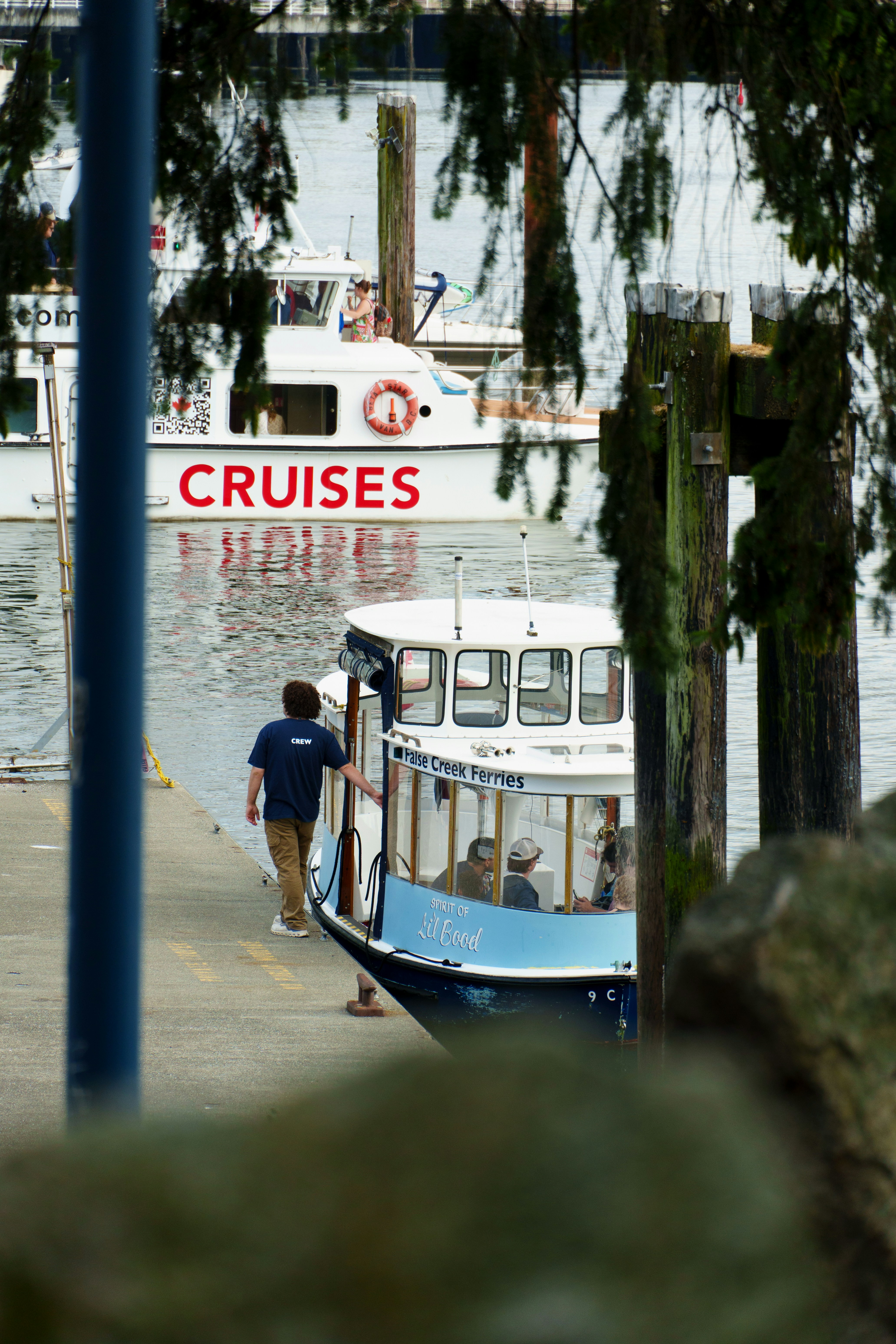 A harbor scene shows a small blue-and-white ferry docked near a larger passenger boat with a bold 'CRUISES' sign, while a worker approaches the vessel at a wooden pier. The image captures a candid moment of maritime activity near the water.