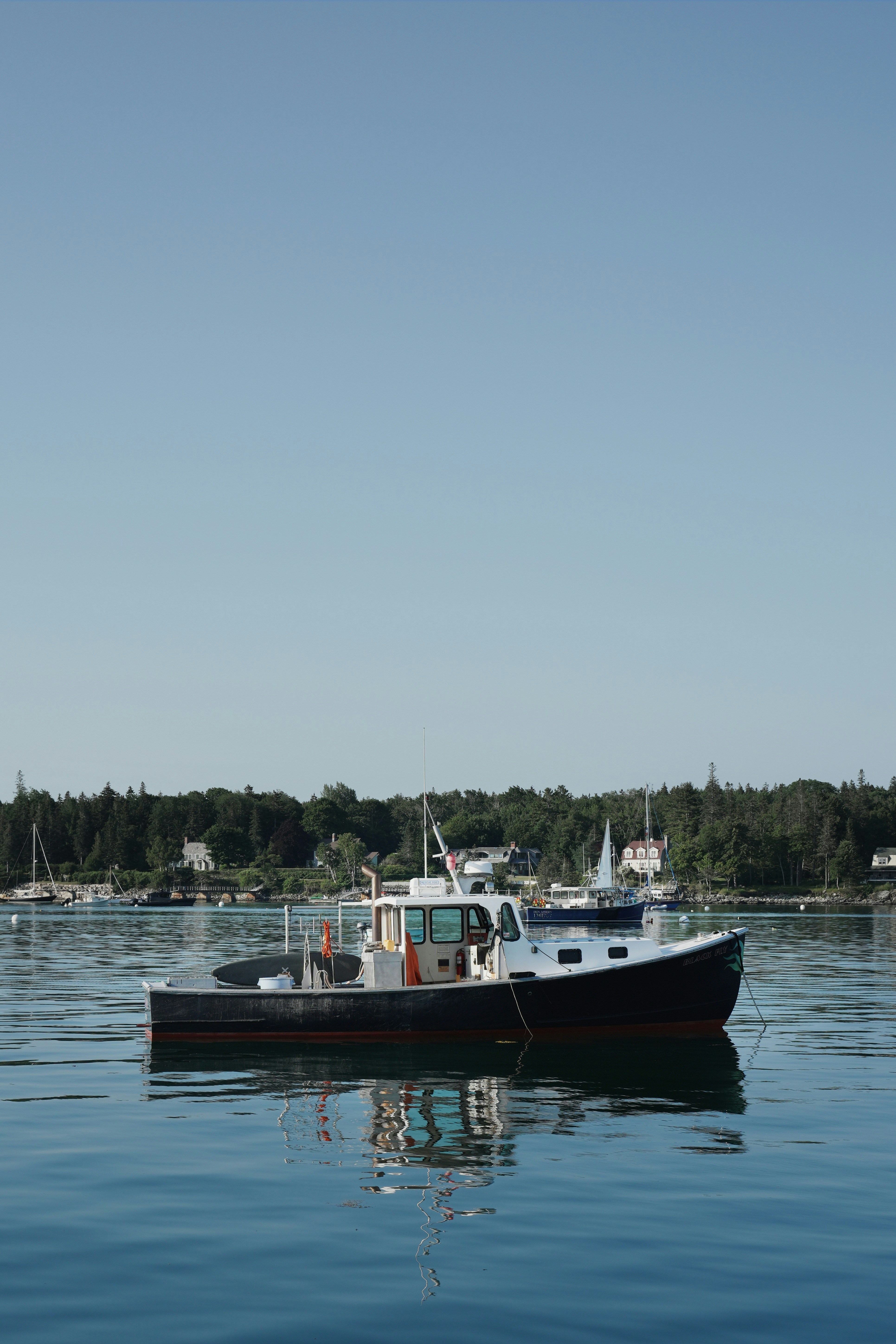 A small fishing boat sits calmly on a glassy harbor with a tree-lined shore and other vessels in the background, its reflection rippling softly in the water. This is a photographic capture of a tranquil harbor scene.