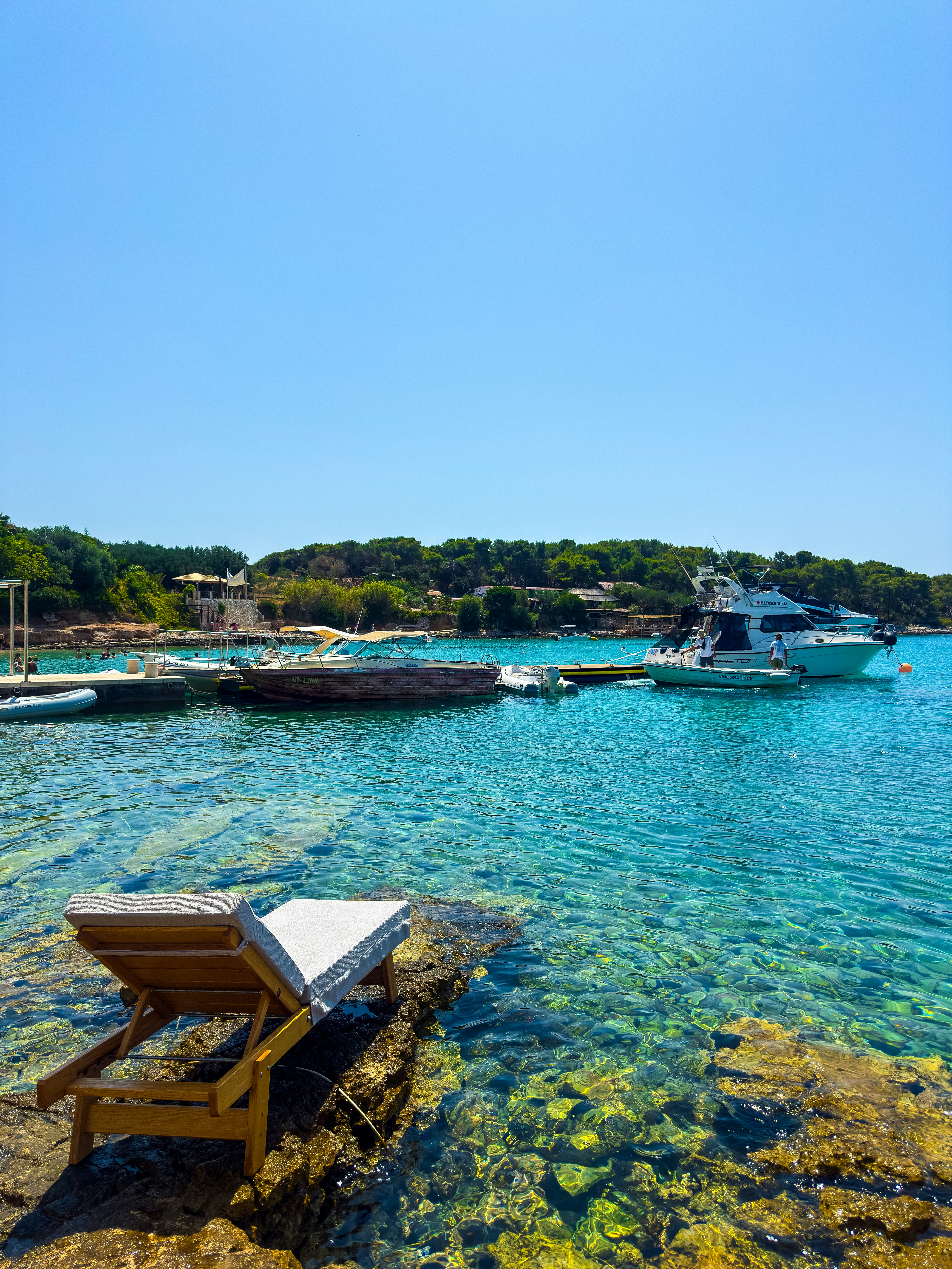 Clear turquoise water with boats at a quiet harbor and a lone lounge chair on rocky shore, inviting a peaceful seaside retreat.