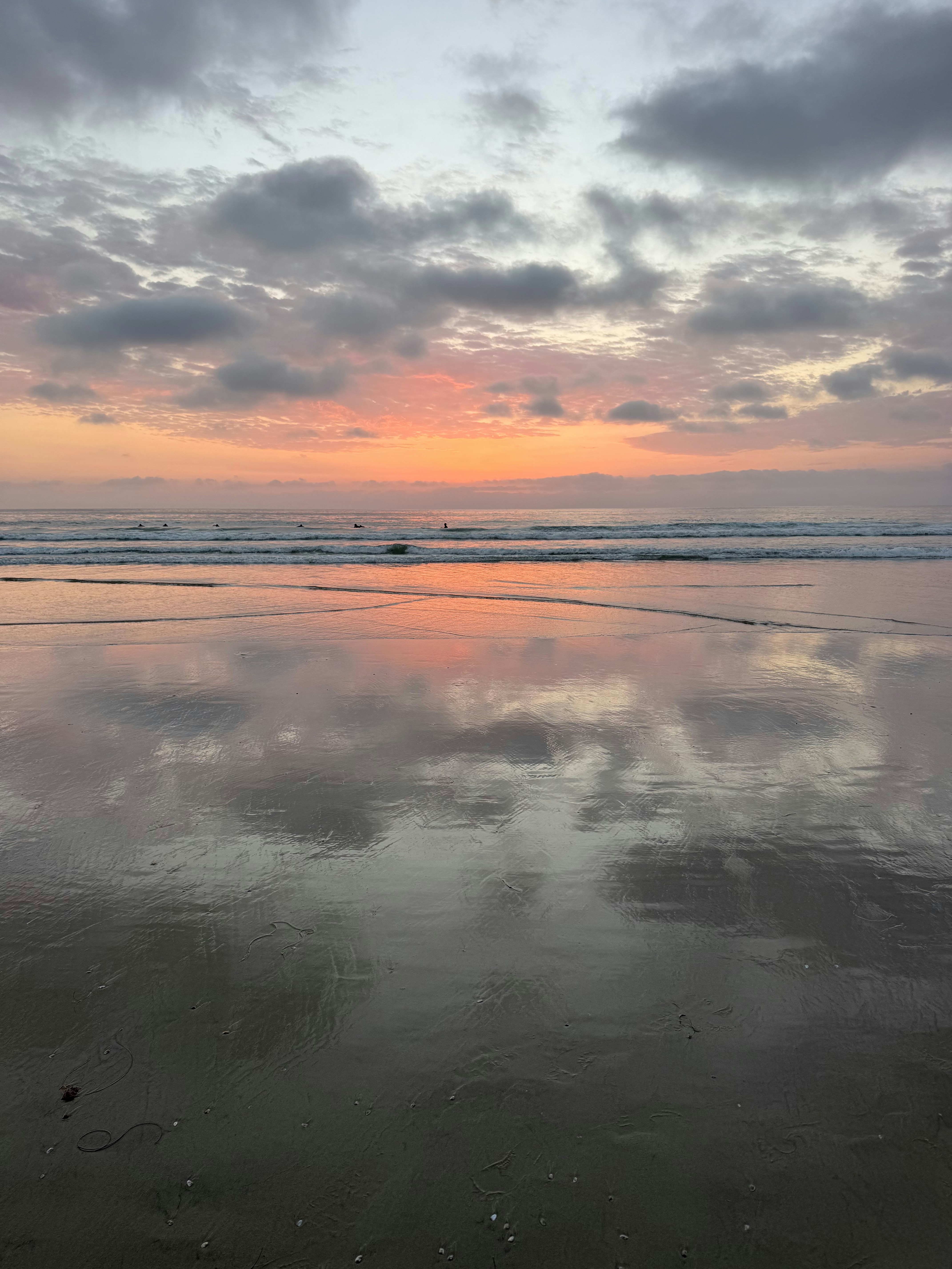 Dusk-lit beach with calm sea and vibrant sky reflecting on wet sand; a tranquil coastal scene without people. This is a photographic capture of the horizon at sunset.