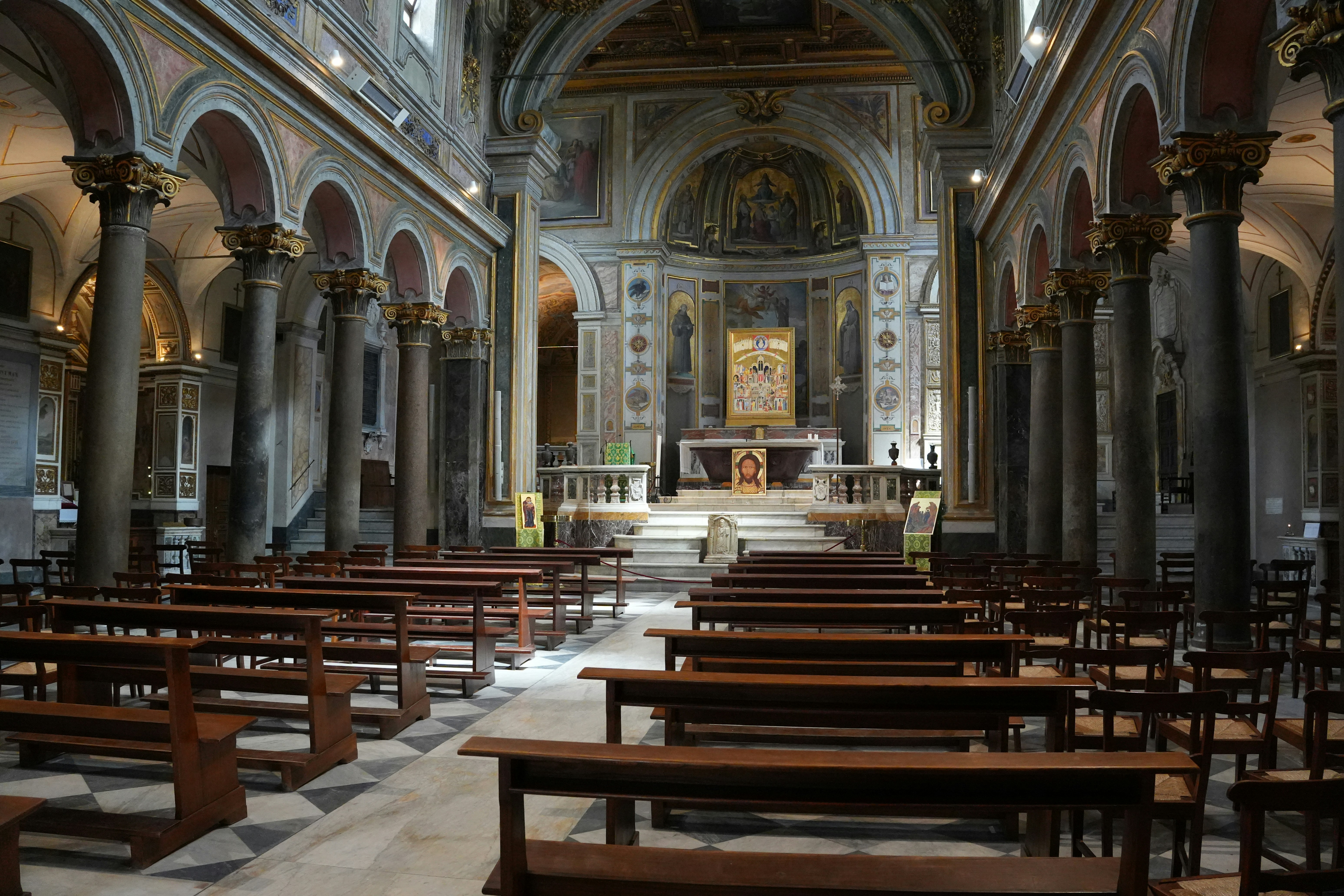 Interior of a historic church with wooden pews leading toward an ornate altar under vaulted ceilings. The space exudes quiet reverence and architectural detail; a painting and arches anchor the focal point of the sanctuary.