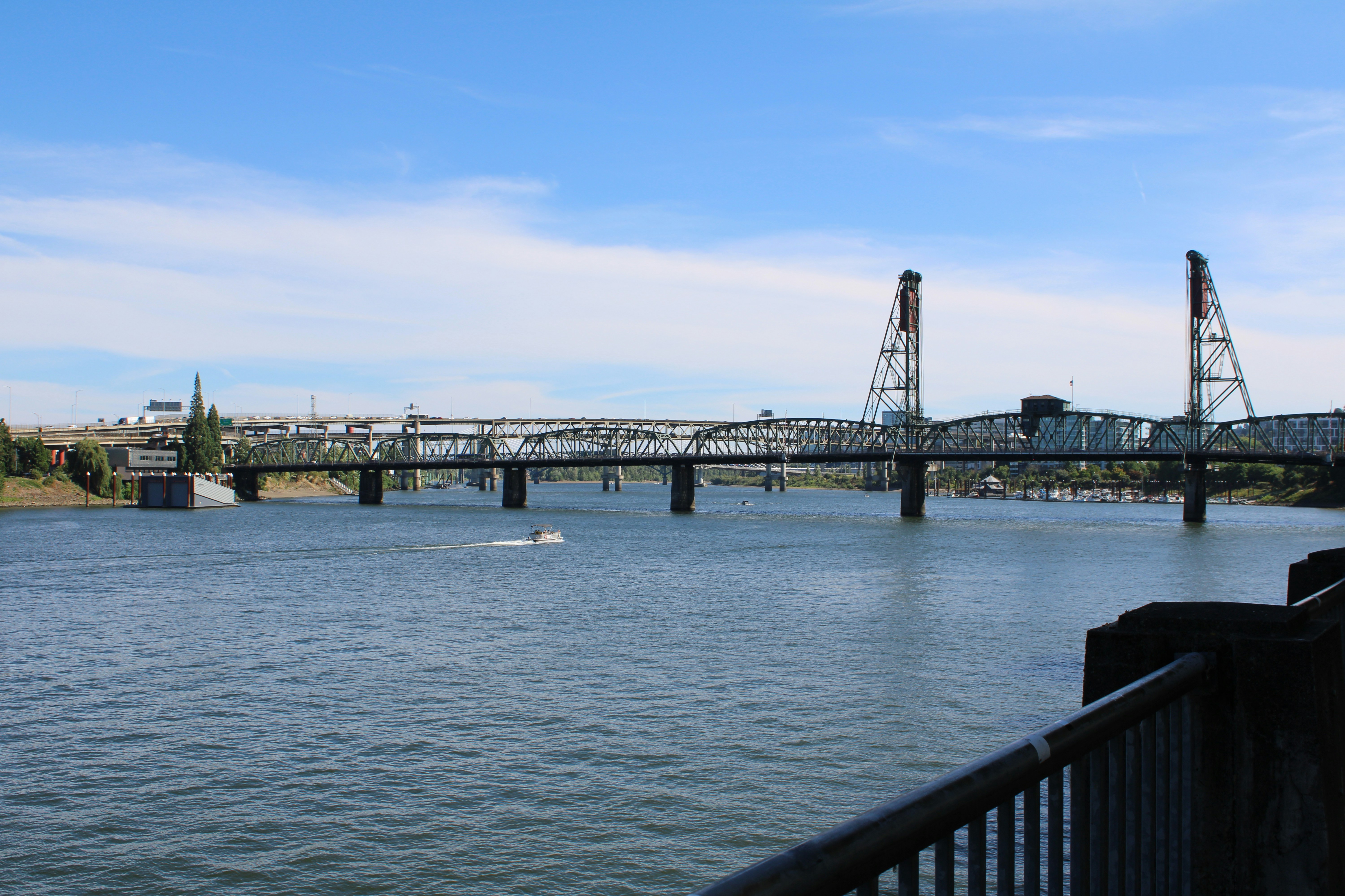 A long steel vertical-lift bridge spans wide river waters under a lightly clouded blue sky as a small boat leaves a wake below.