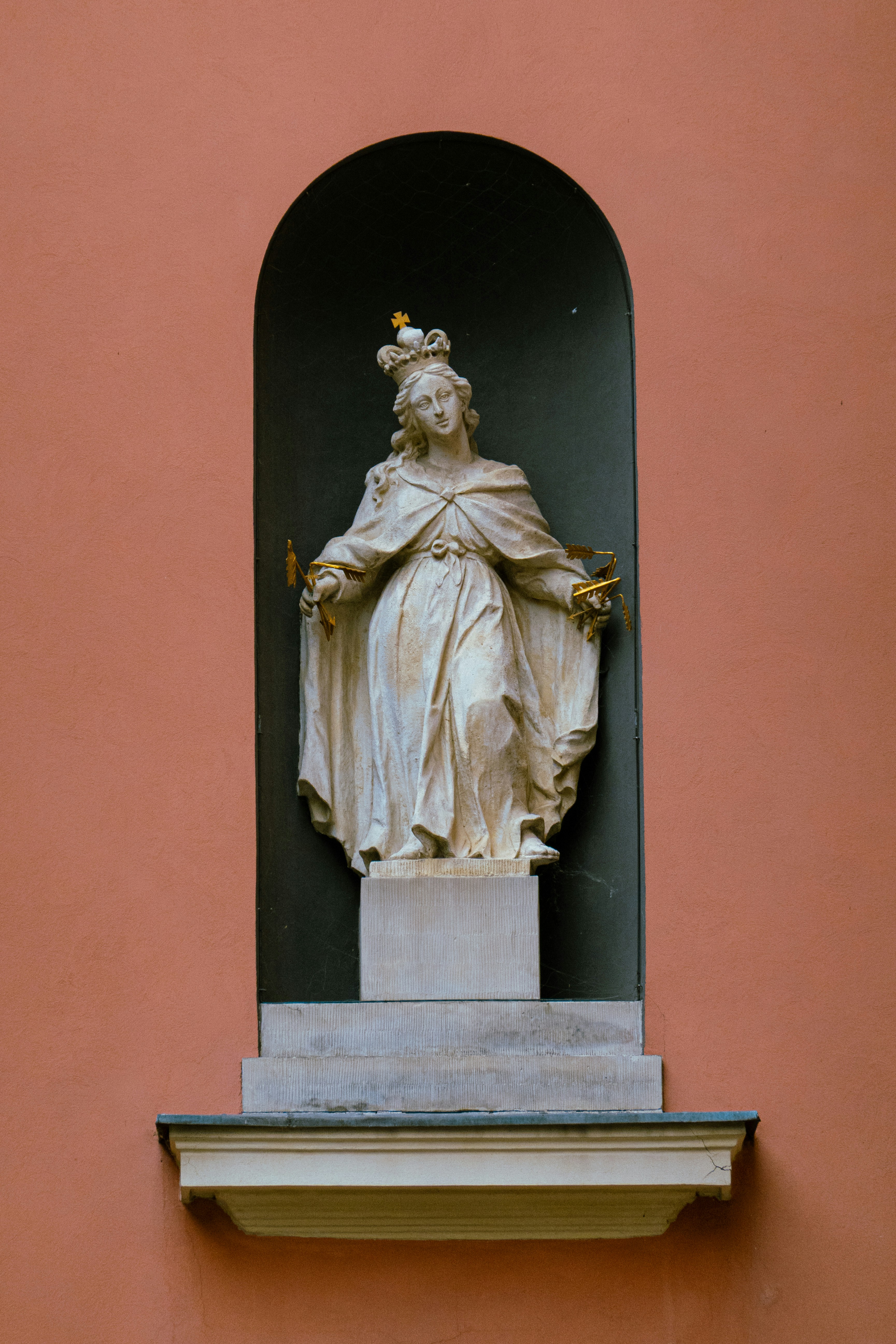 Stone statue in a niche with a dark recessed center, set against a warm terracotta wall; pedestal and architectural ledge visible.