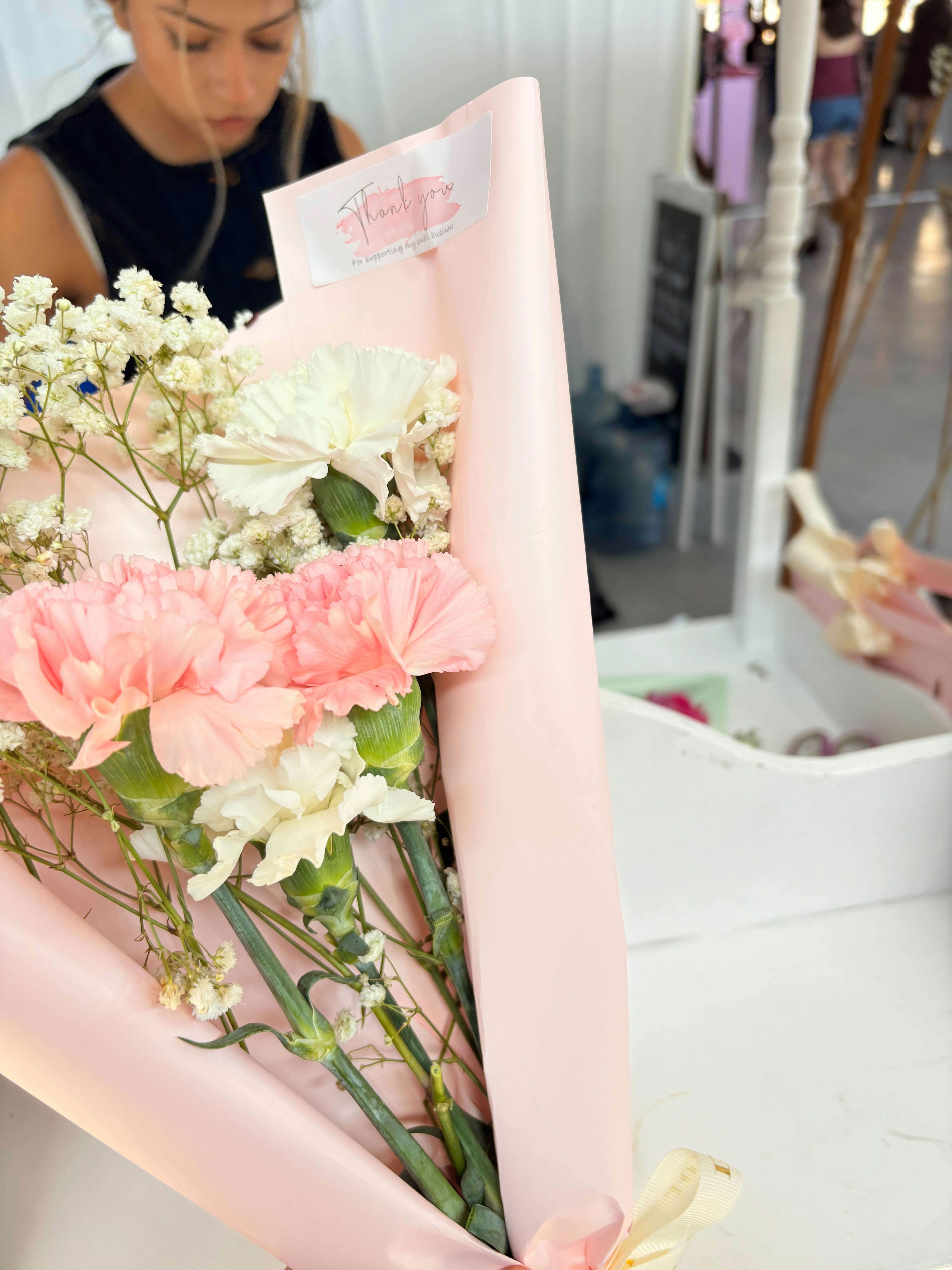 Soft pink bouquet wrapped in pastel paper with delicate white flowers, held against a blurred background at a floral display.