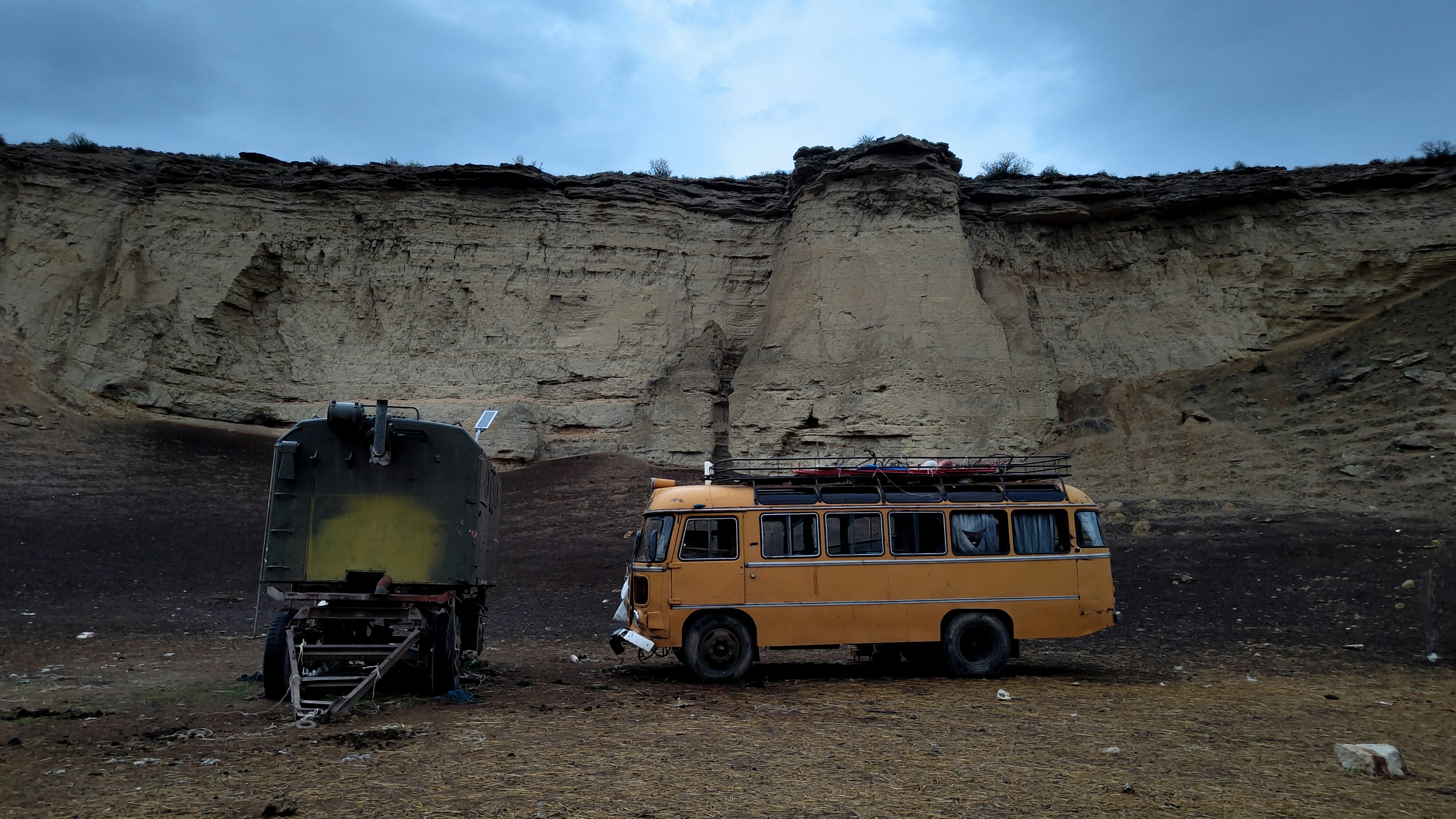 Yellow bus rests beside a weathered tanker at the base of eroded cliffs, with a moody sky overhead.
