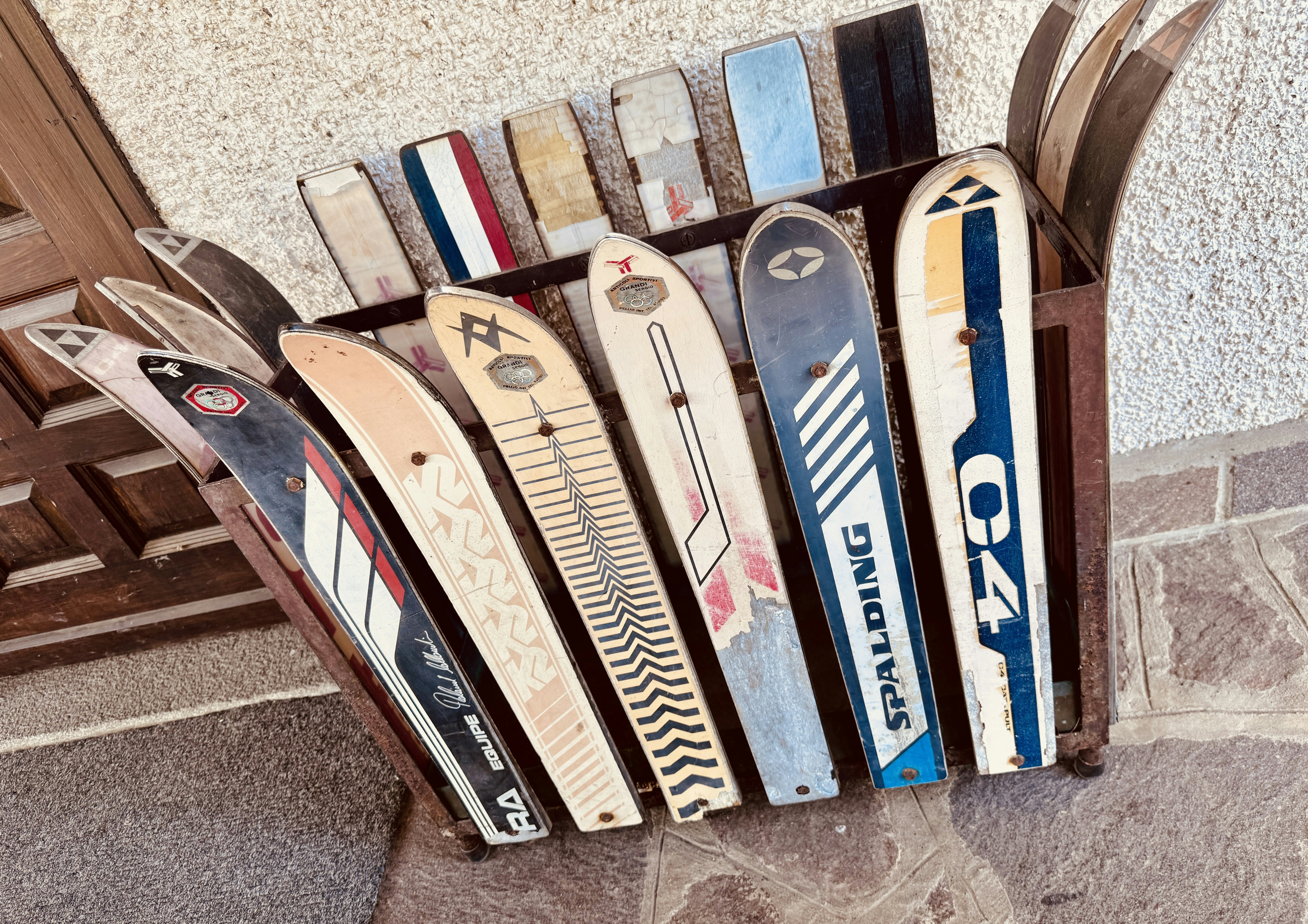 Row of old inline skates with scuffed surfaces and faded paint stacked along a wall; the scene captures nostalgic athletic gear in a rustic setting.