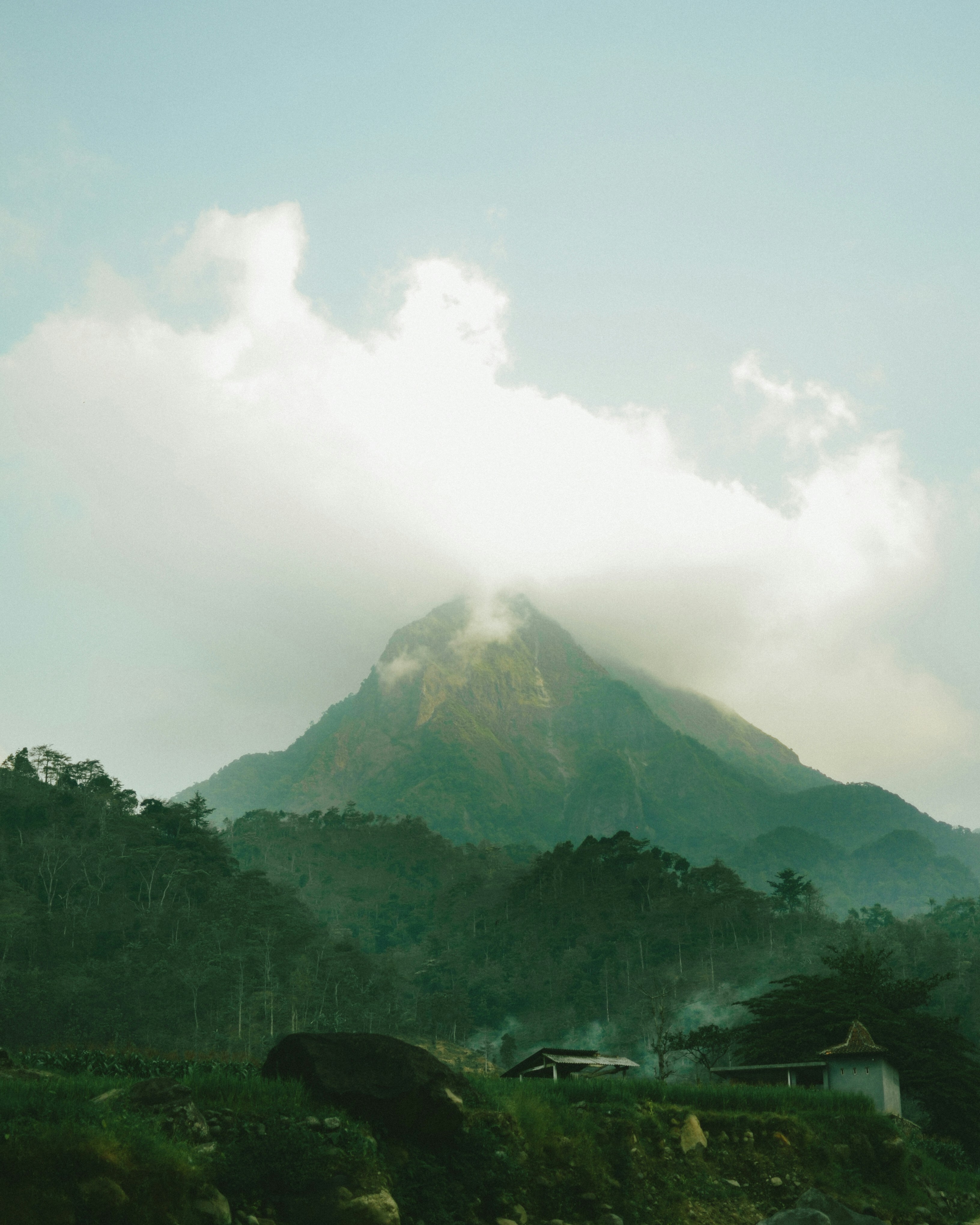 Misty mountain peak rises above a forested valley with a small settlement at the foothills, clouds resting over summit in soft morning light.