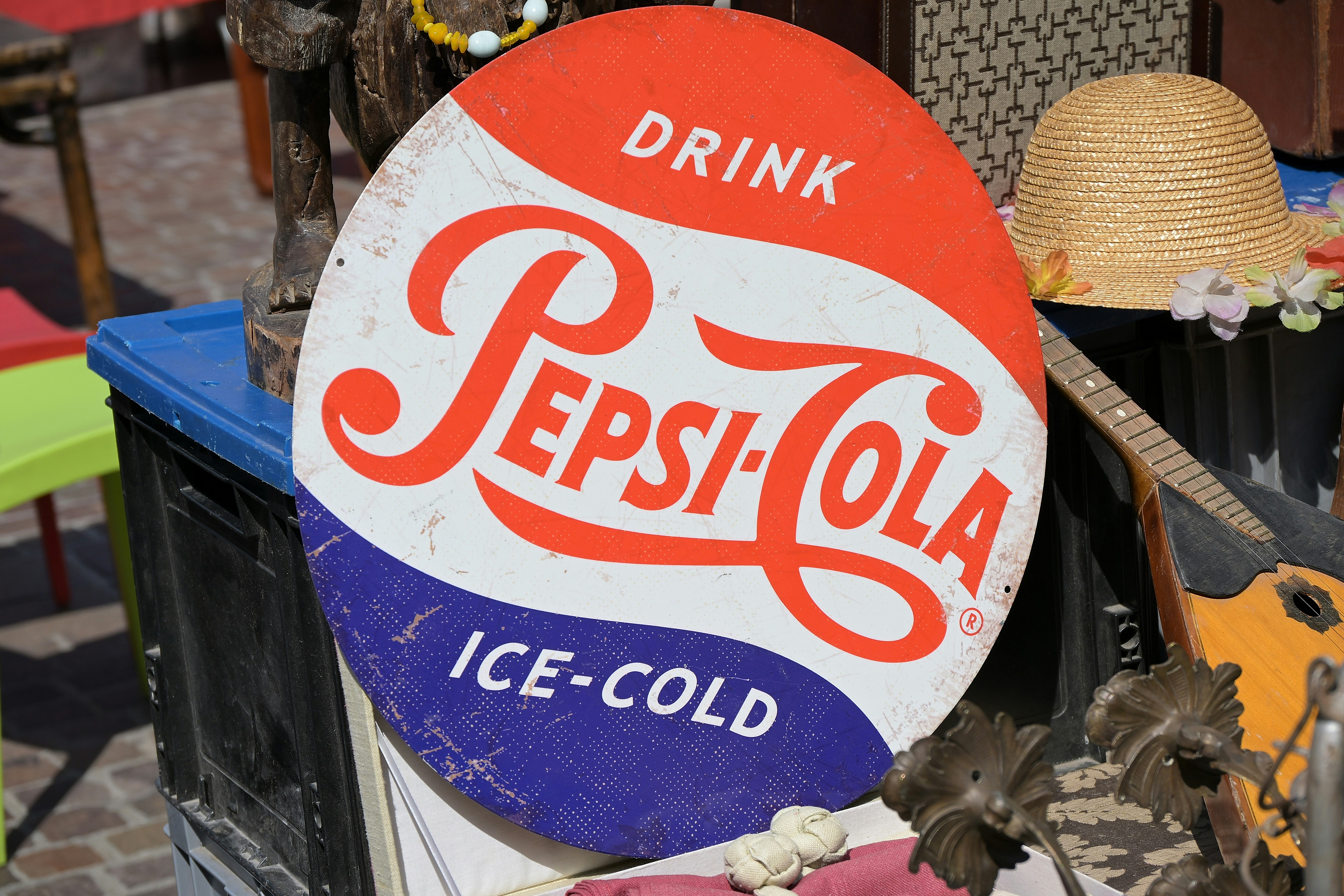 A vintage circular Pepsi-Cola sign leaning among market clutter, with hats and decorative items in the background.
