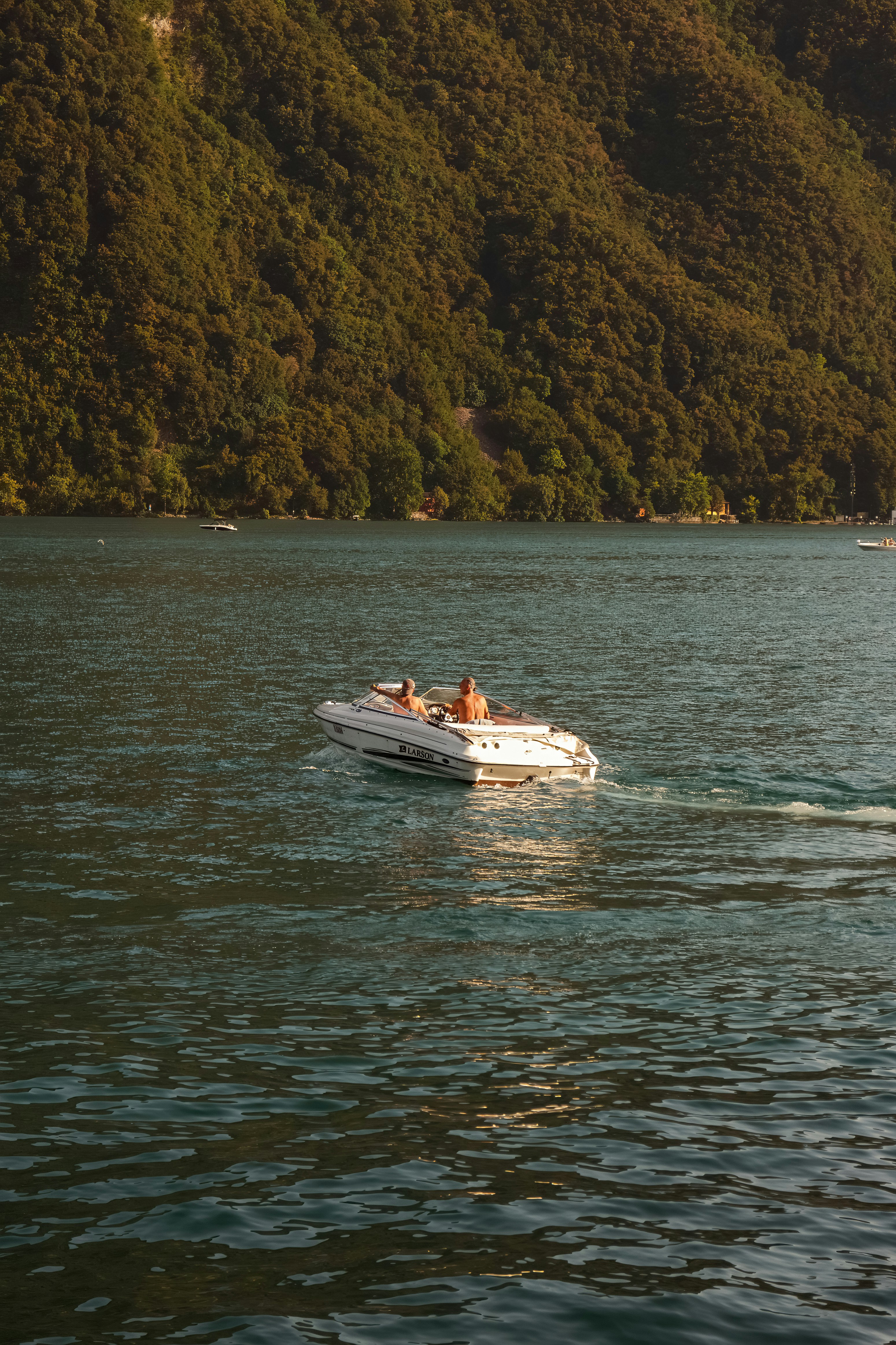 White motorboat glides across a calm lake with a forested hillside in the background, two occupants enjoying the ride. Coastal scenery with gentle ripples; suitable for a travel or nature scene.