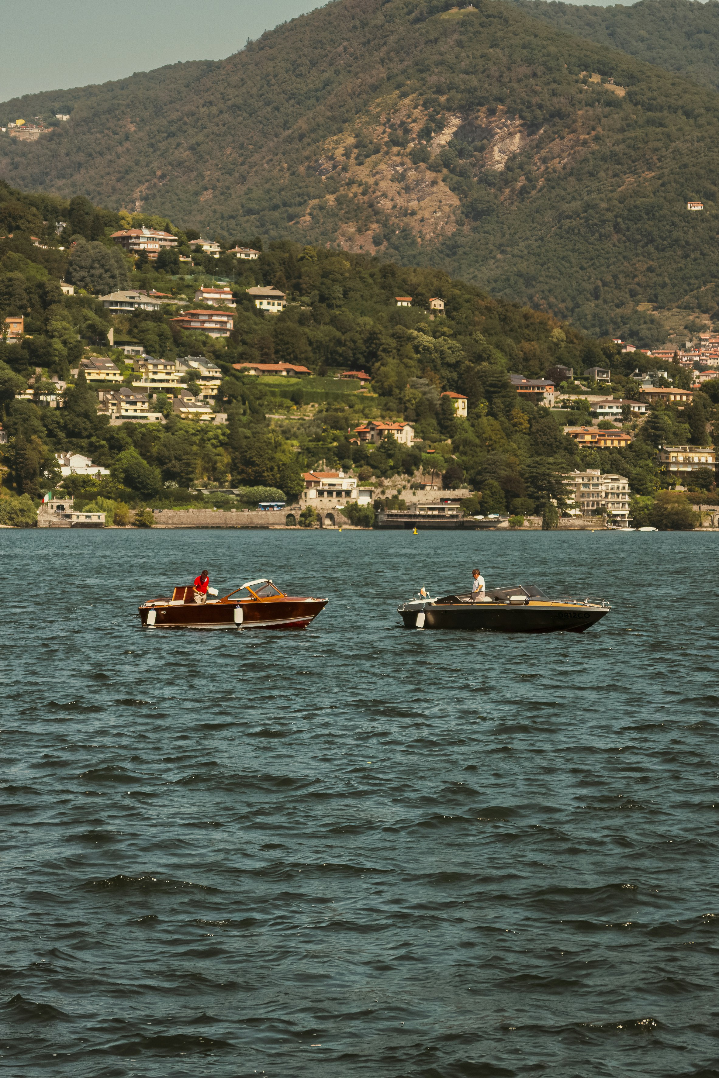 Two motorboats drift on a calm lake with a hillside village and mountains in the background.