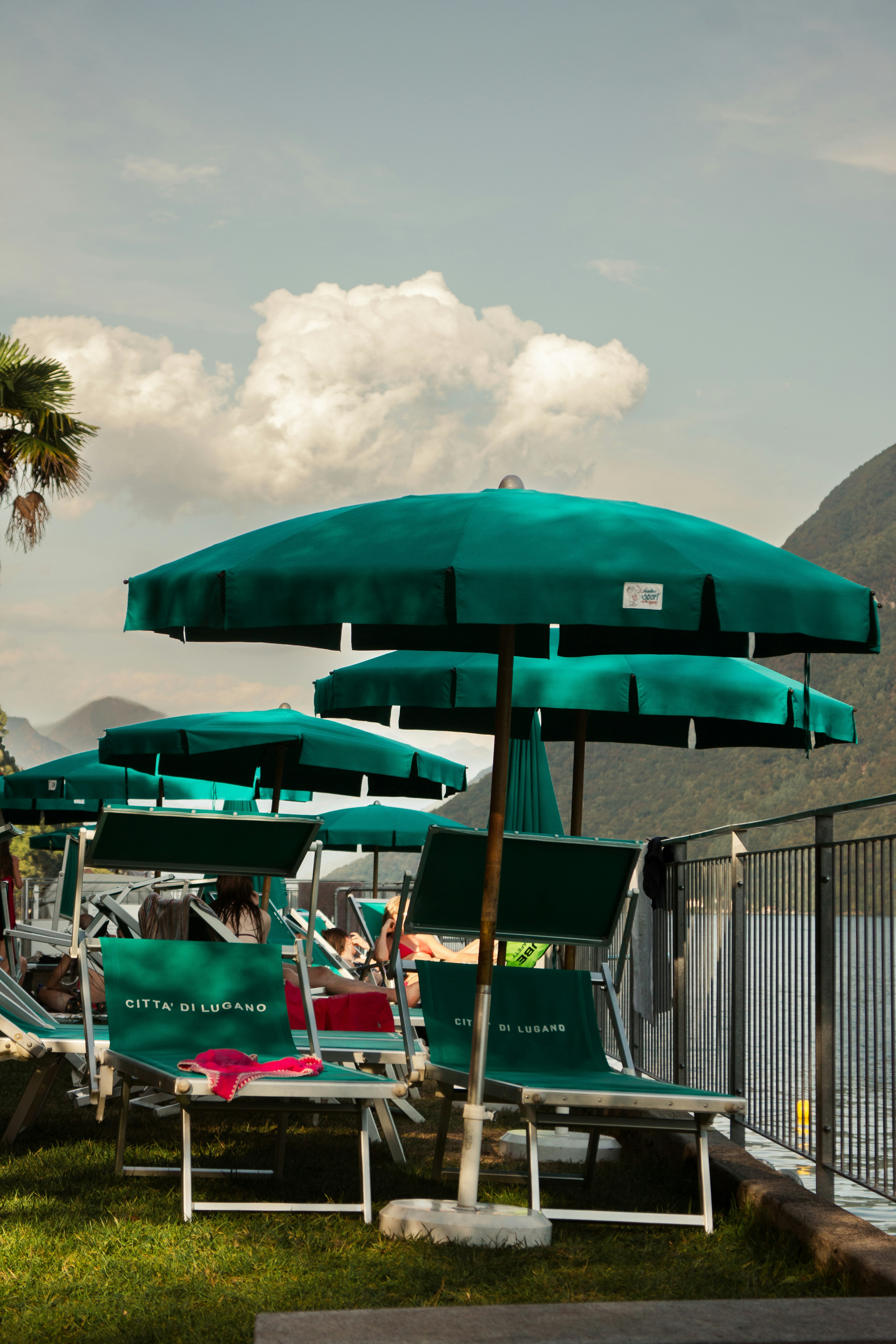 Row of turquoise beach umbrellas and lounge chairs along a railing by the water, with distant mountains and a bright sky in the background. This is a staged outdoor resort scene with signage on chairs indicating a city coast setting.