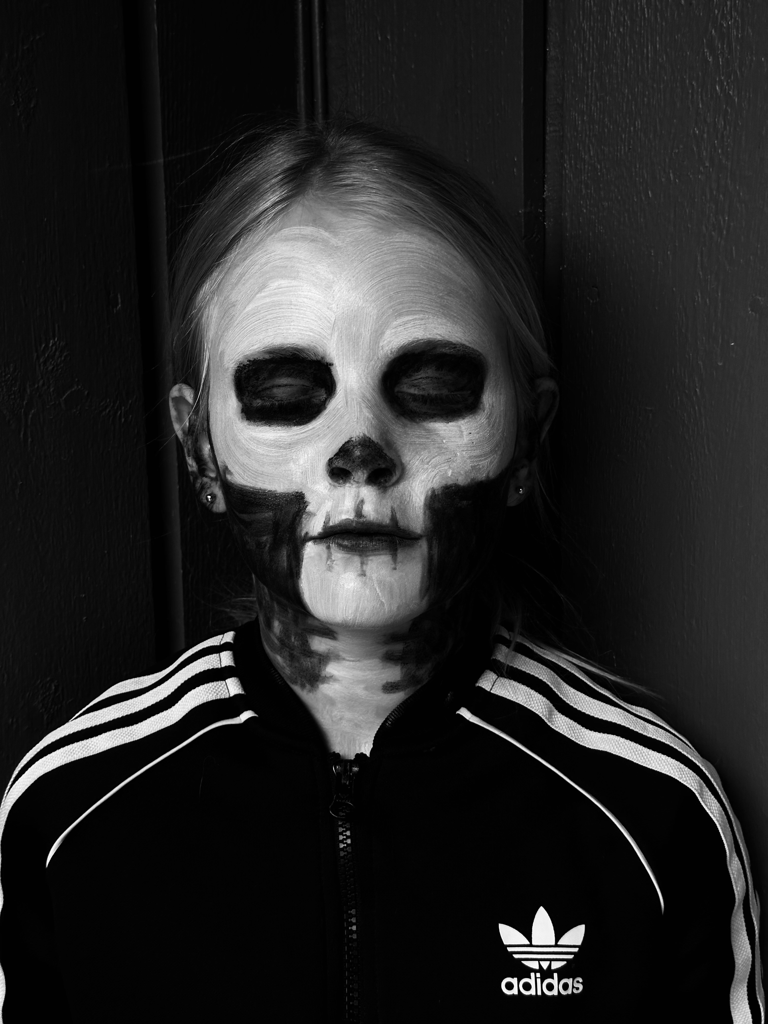 A young girl with closed eyes and face paint in front of dark wall. Face is painted as a scull.