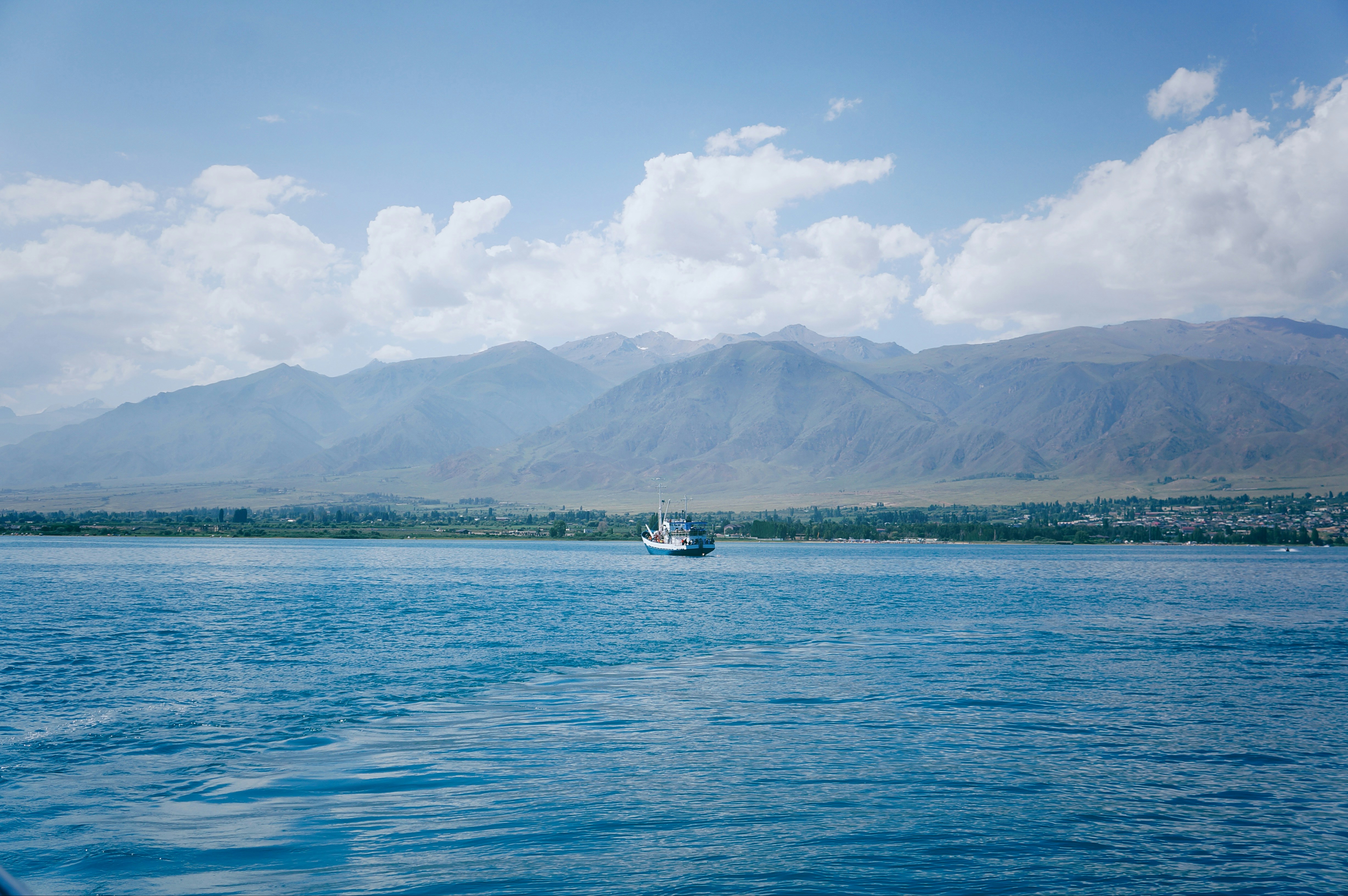 Small boat crosses calm blue water with a mountain range in the distance and a bright sky above; a tranquil seascape with a single focal vessel. This is a landscape photograph.