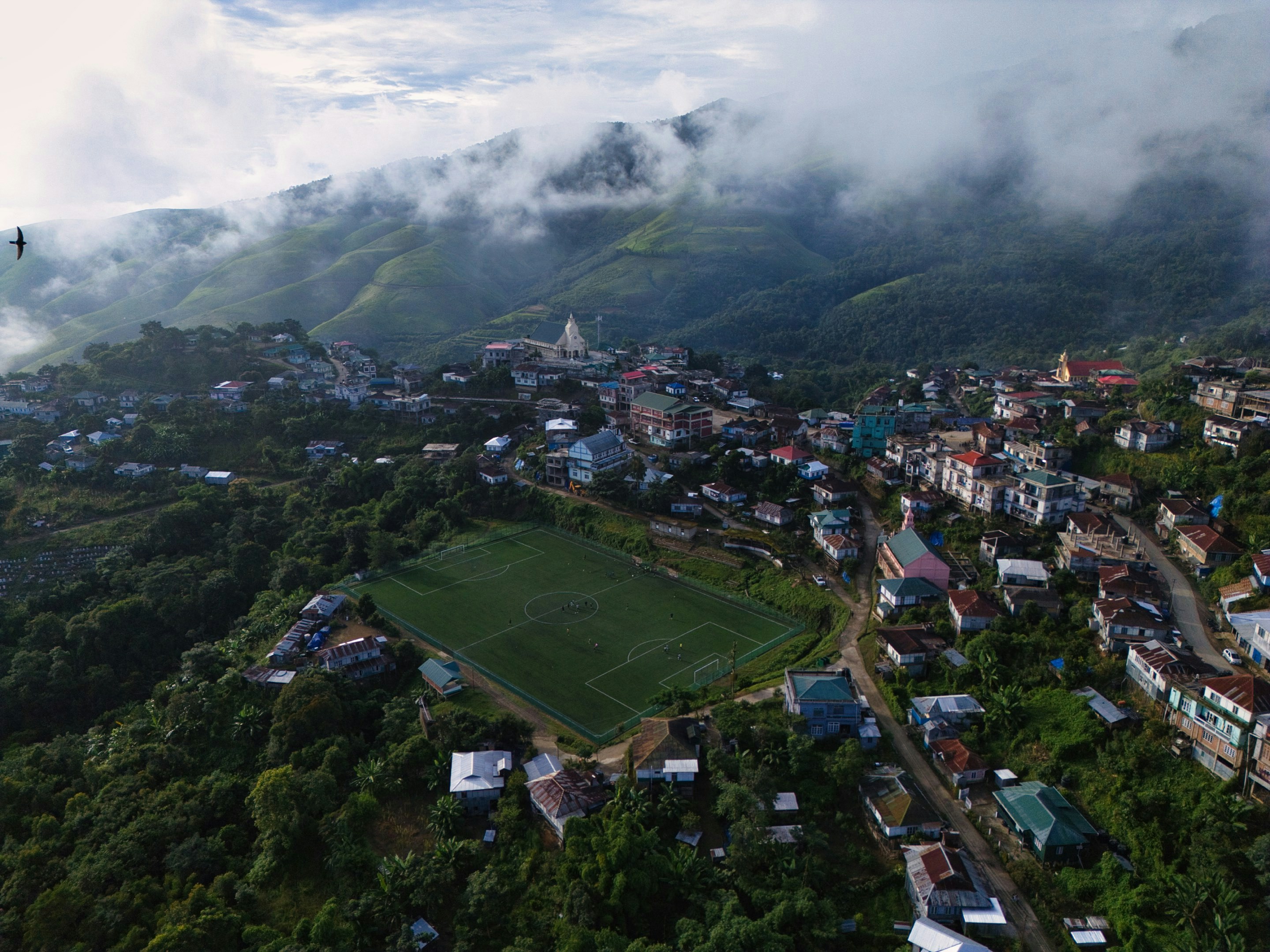 A drone view reveals a small town clustered around a green football field, with misty mountains in the background and a network of colorful houses dotting the hillside.