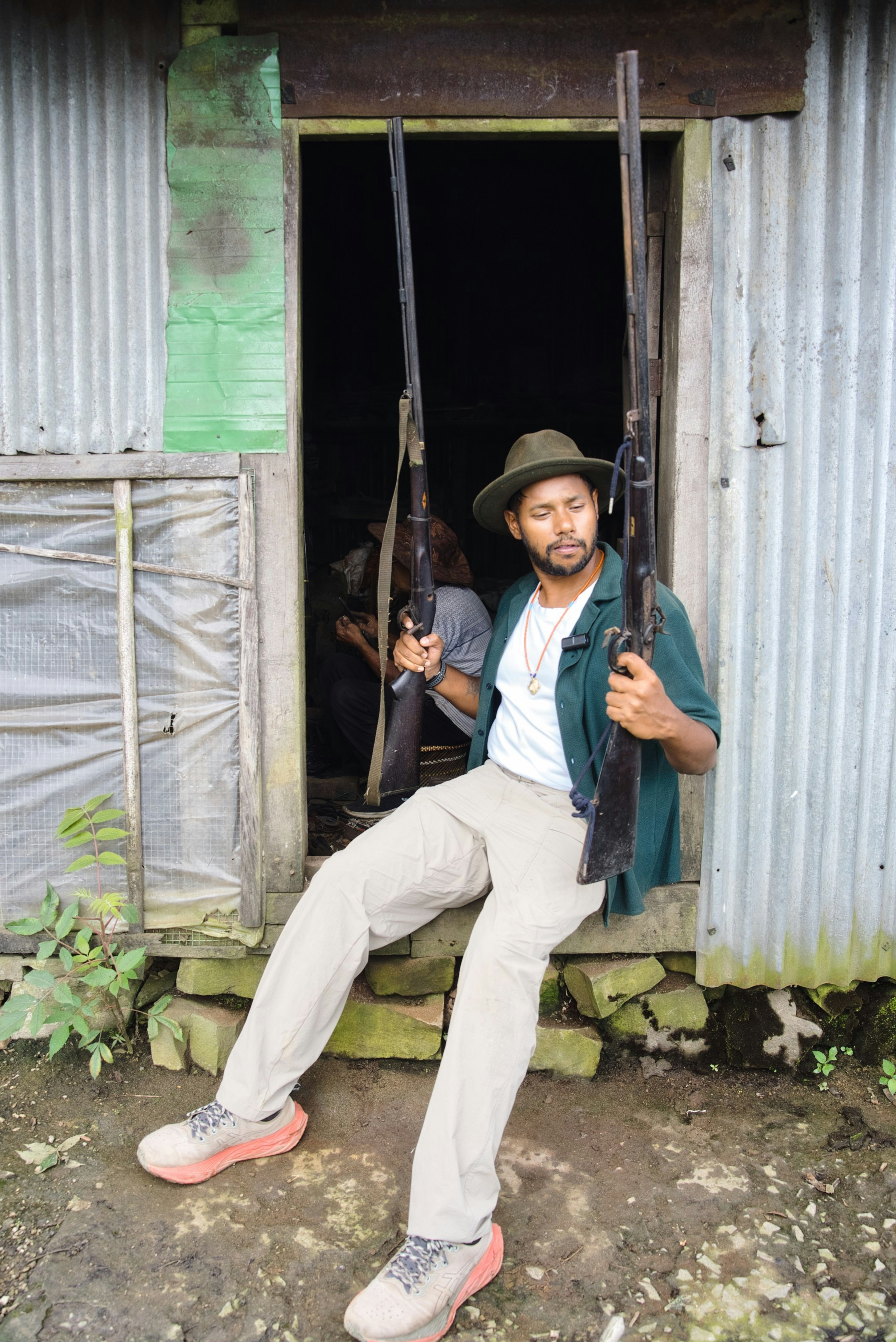 A man with two rifles sits in a doorway.