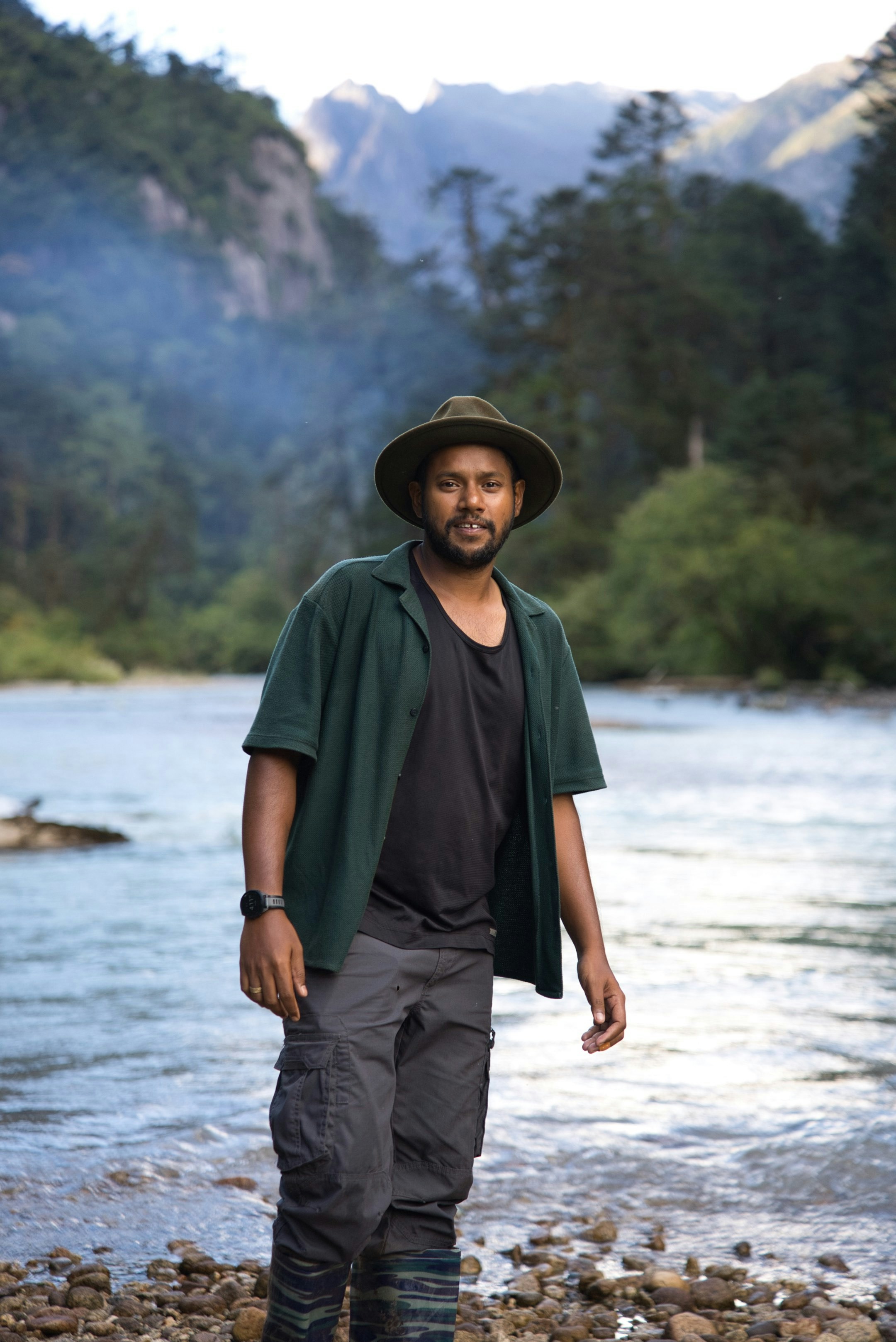 A man stands on a rocky riverbank with a calm river and forested mountains in the background, wearing a green shirt and hat. A subtle plume of mist rises as the scene captures outdoor exploration.