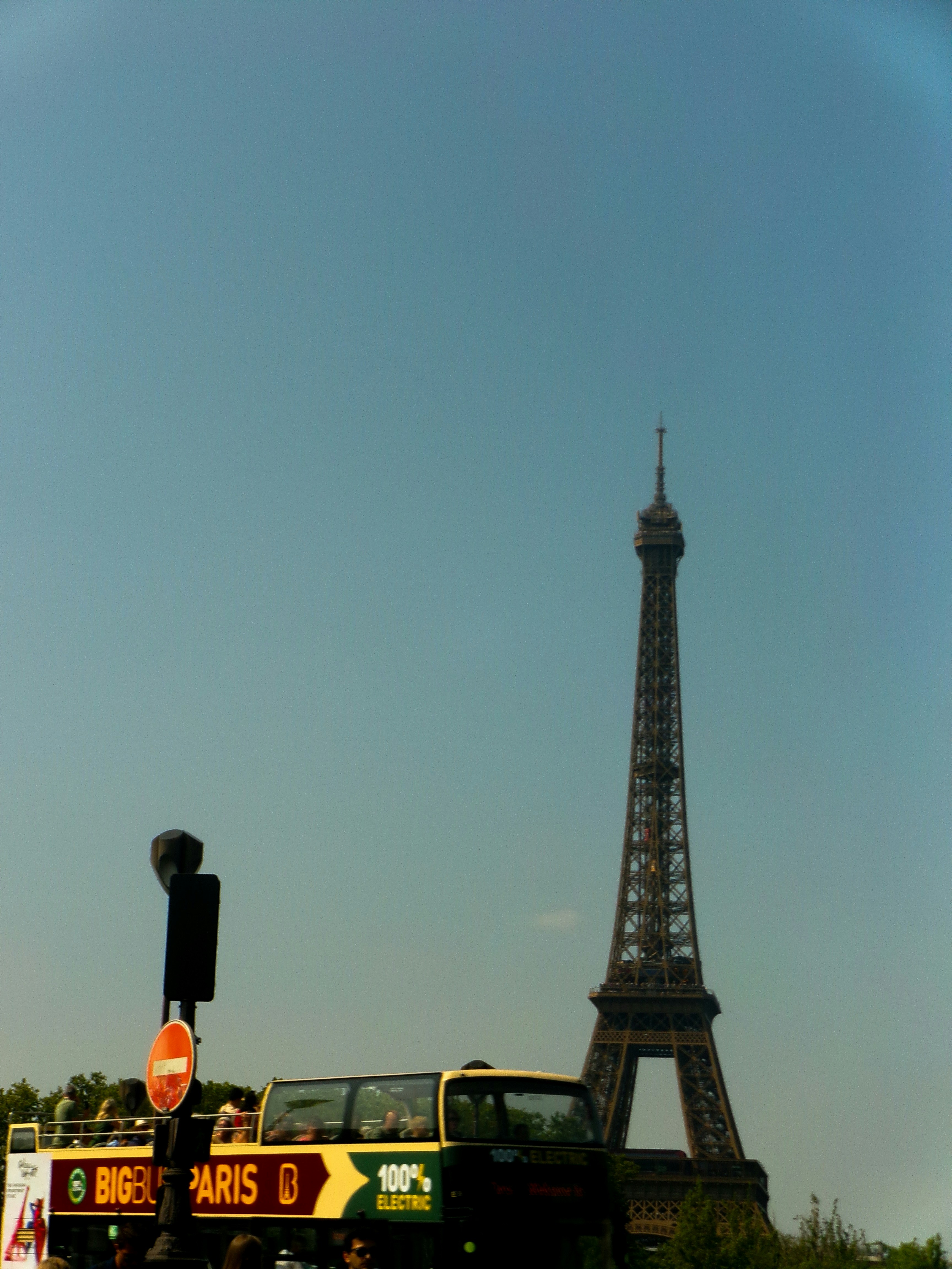 A tour bus and urban signage sit at the base of the Eiffel Tower against a clear sky; the scene captures a bustling, sunny moment in Paris with a hint of motion from passing vehicles. (Note: this is a photograph, not a painting or illustration.)