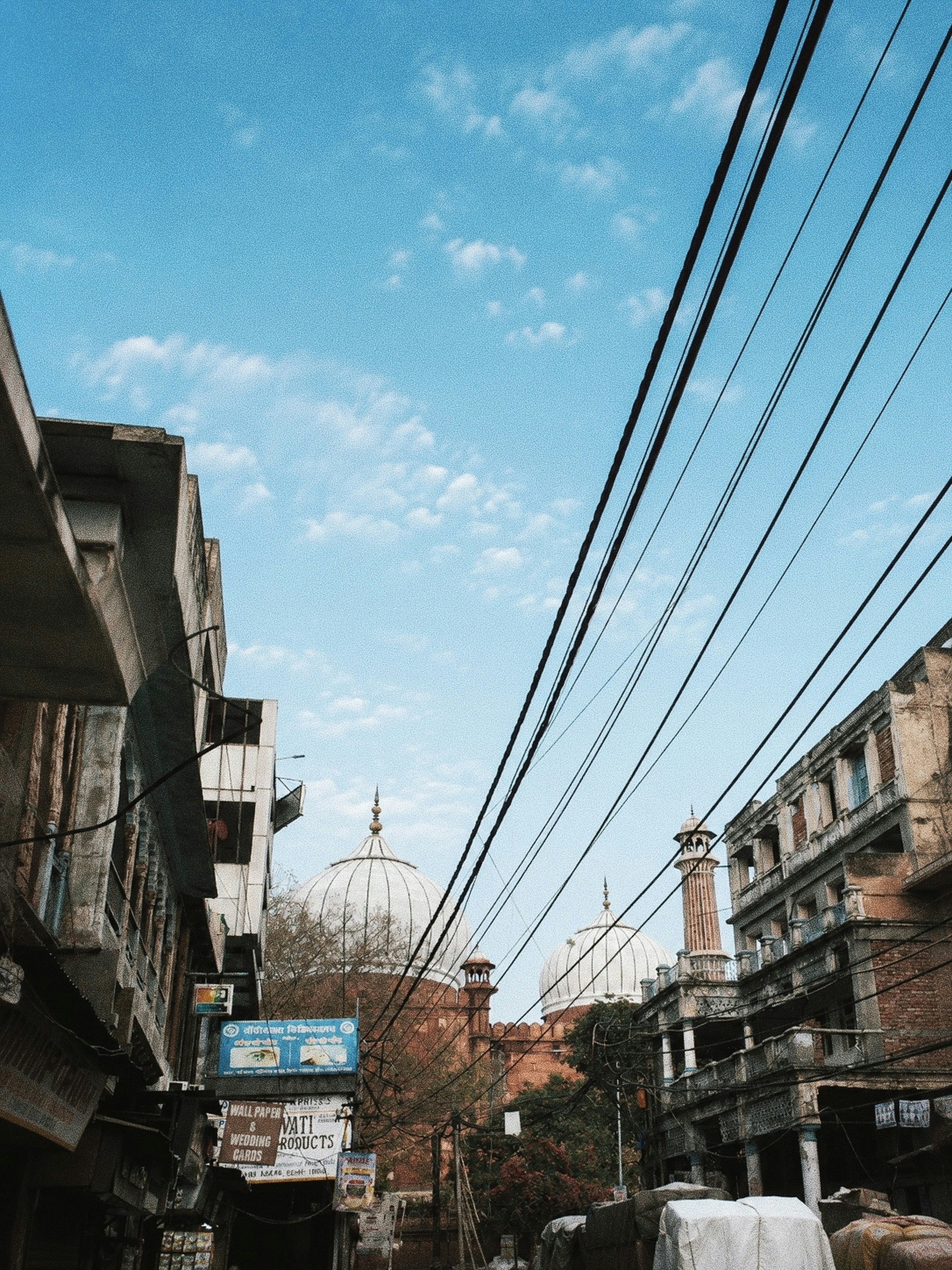 City street with domed mosque silhouettes peeking above buildings, electric wires crossing the frame, blue sky. Photograph of a dense urban landscape with a hint of cultural architecture.