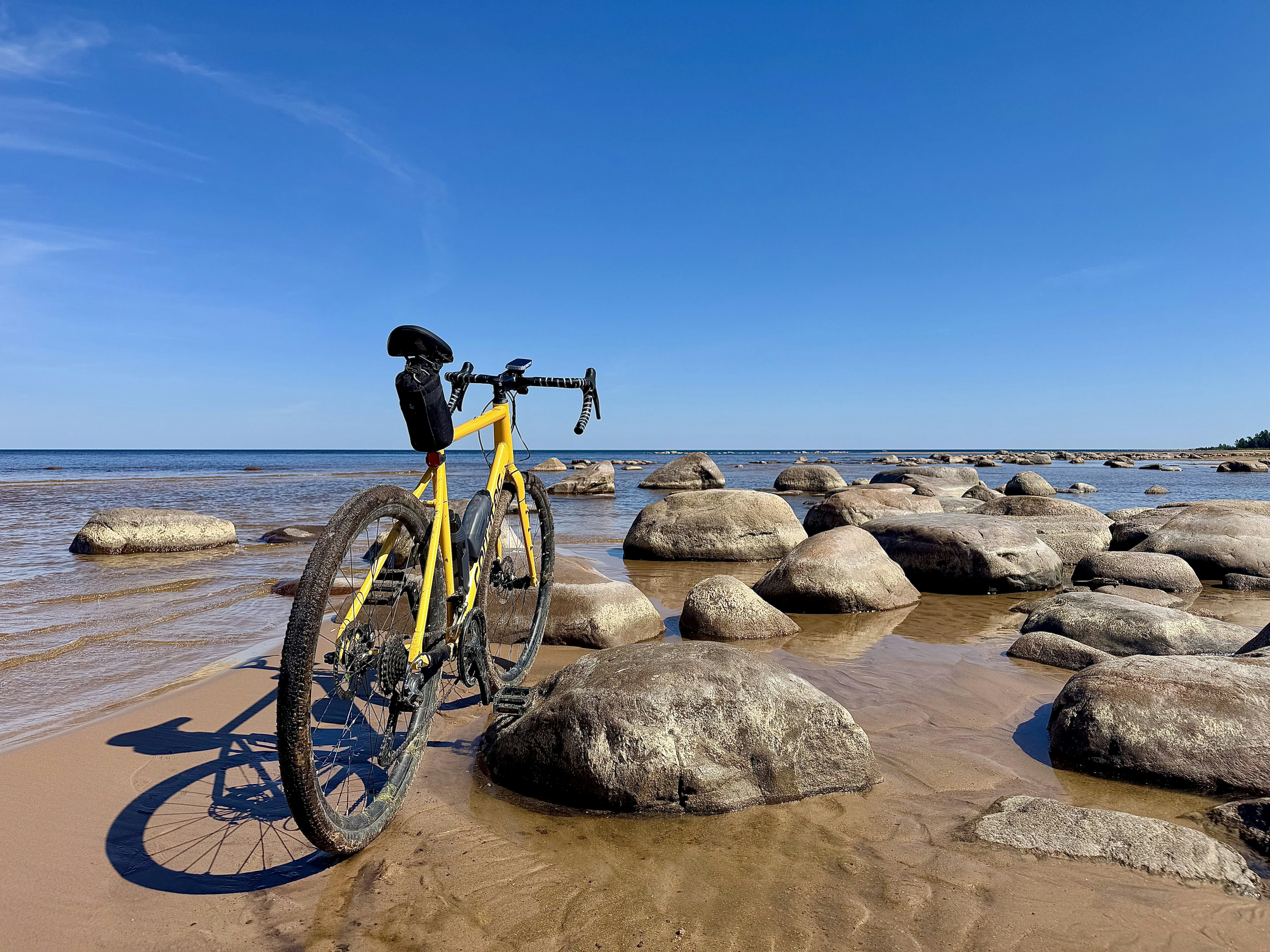 Bright yellow bike resting on wet sand with smooth stones and calm sea under a blue sky.