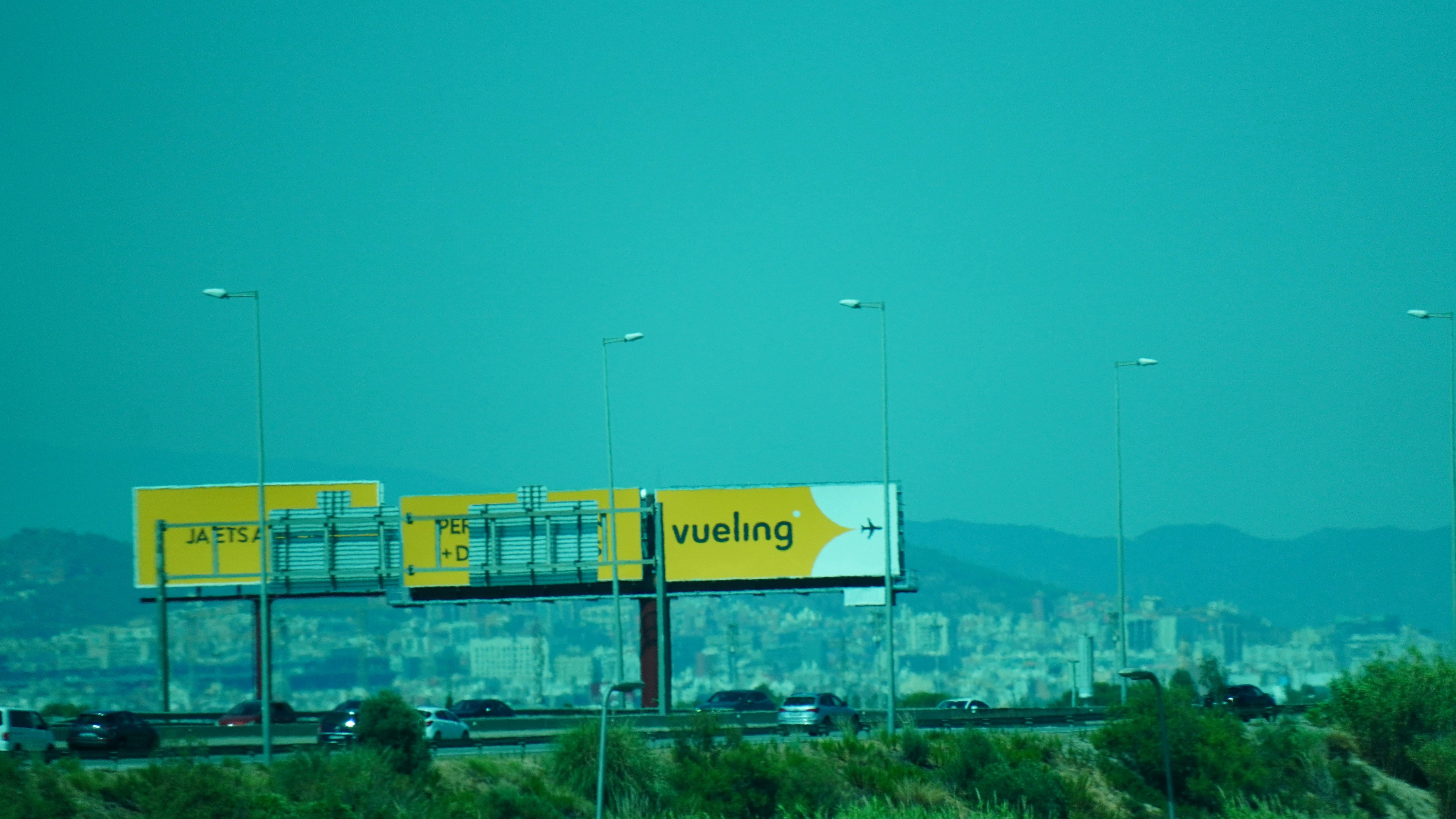Wide highway scene with distant cityscape and bold yellow billboards; foreground shows vegetation and passing cars, suggesting travel and movement. A digital-like blue-green color cast dominates, indicating a stylized or post-processed landscape.